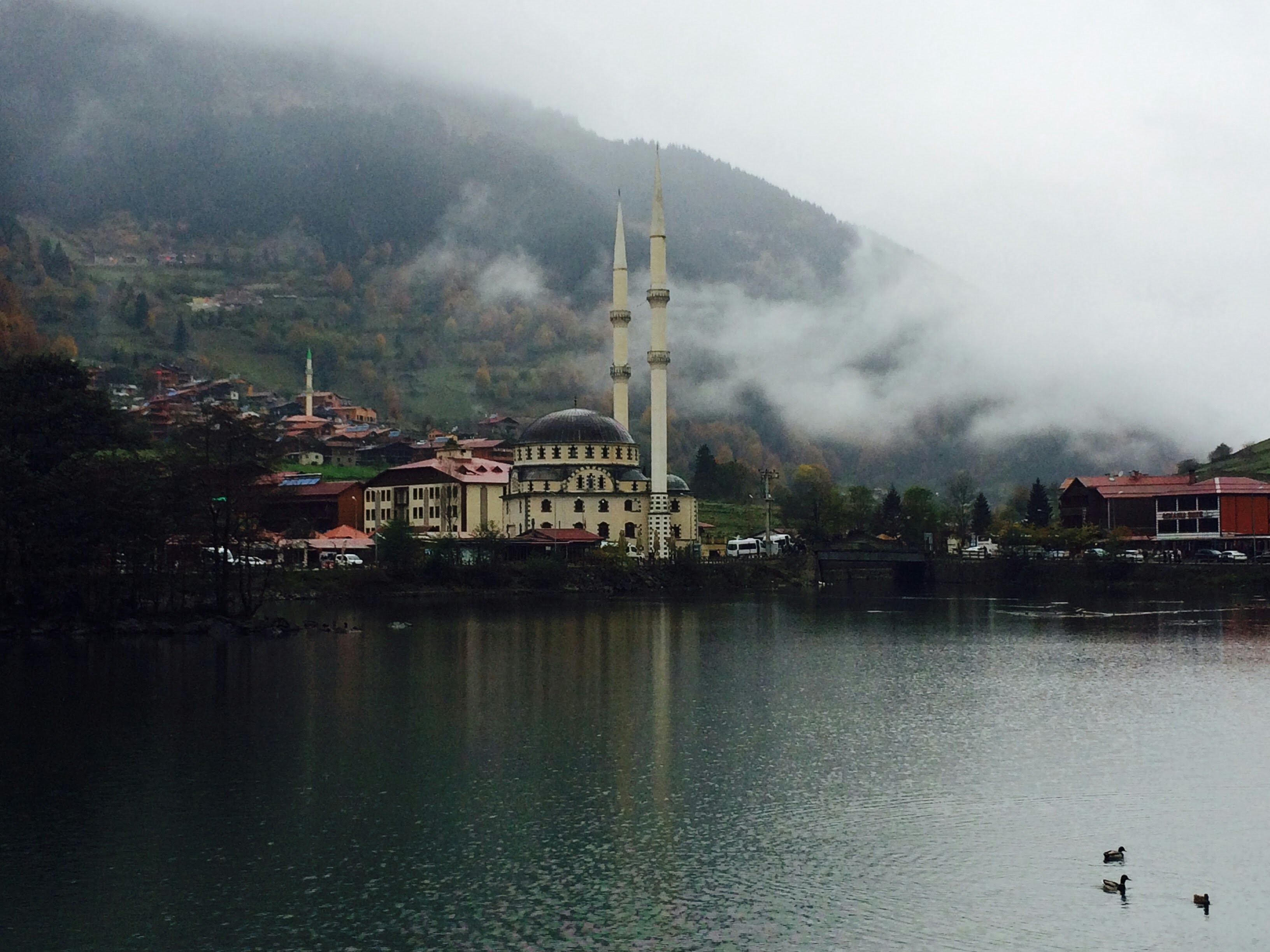 Mosque with twin minarets by a serene lake, surrounded by misty hills and overcast skies.