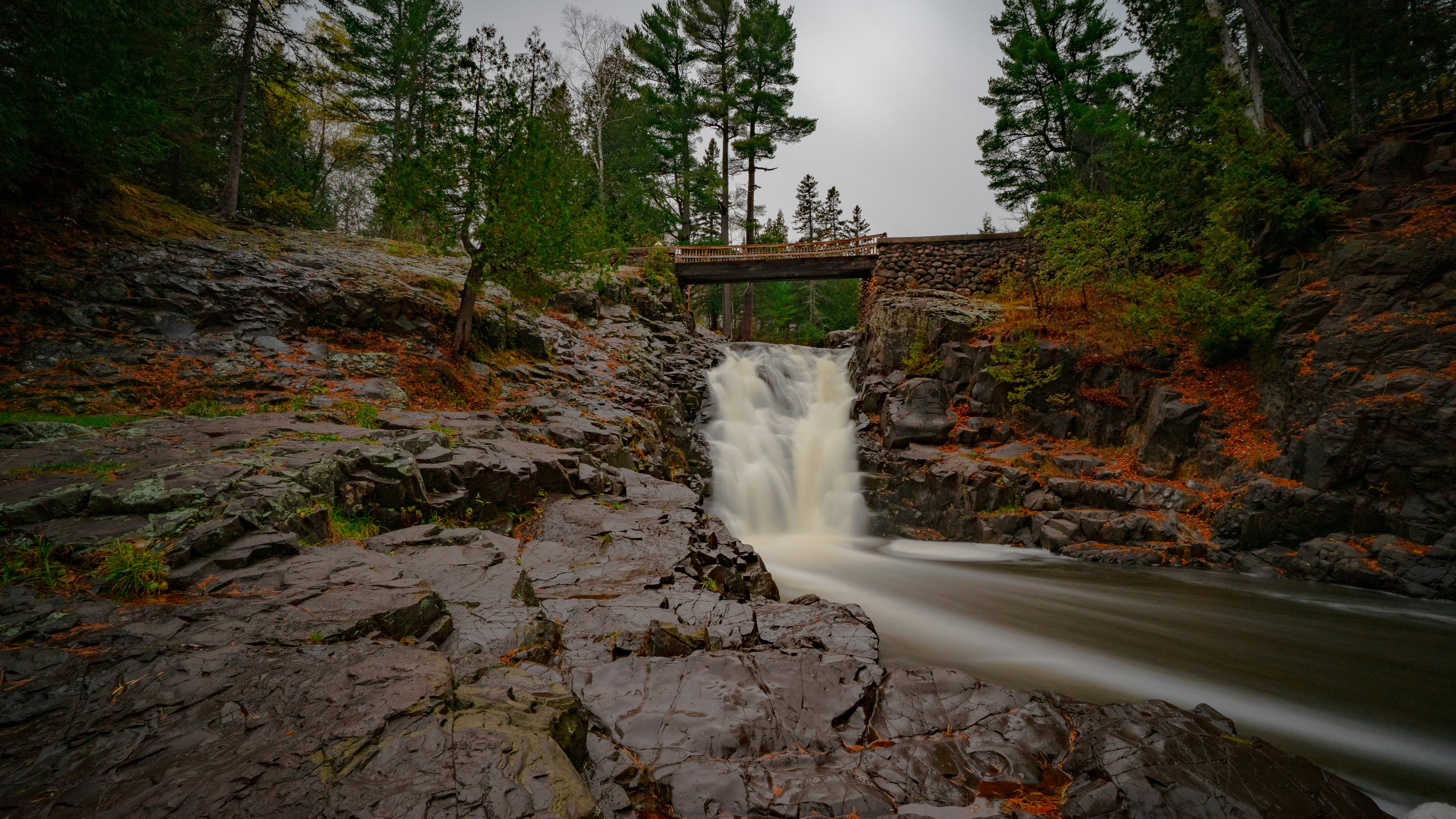 Waterfall flowing through rocky terrain with a wooden bridge above, surrounded by autumn foliage.