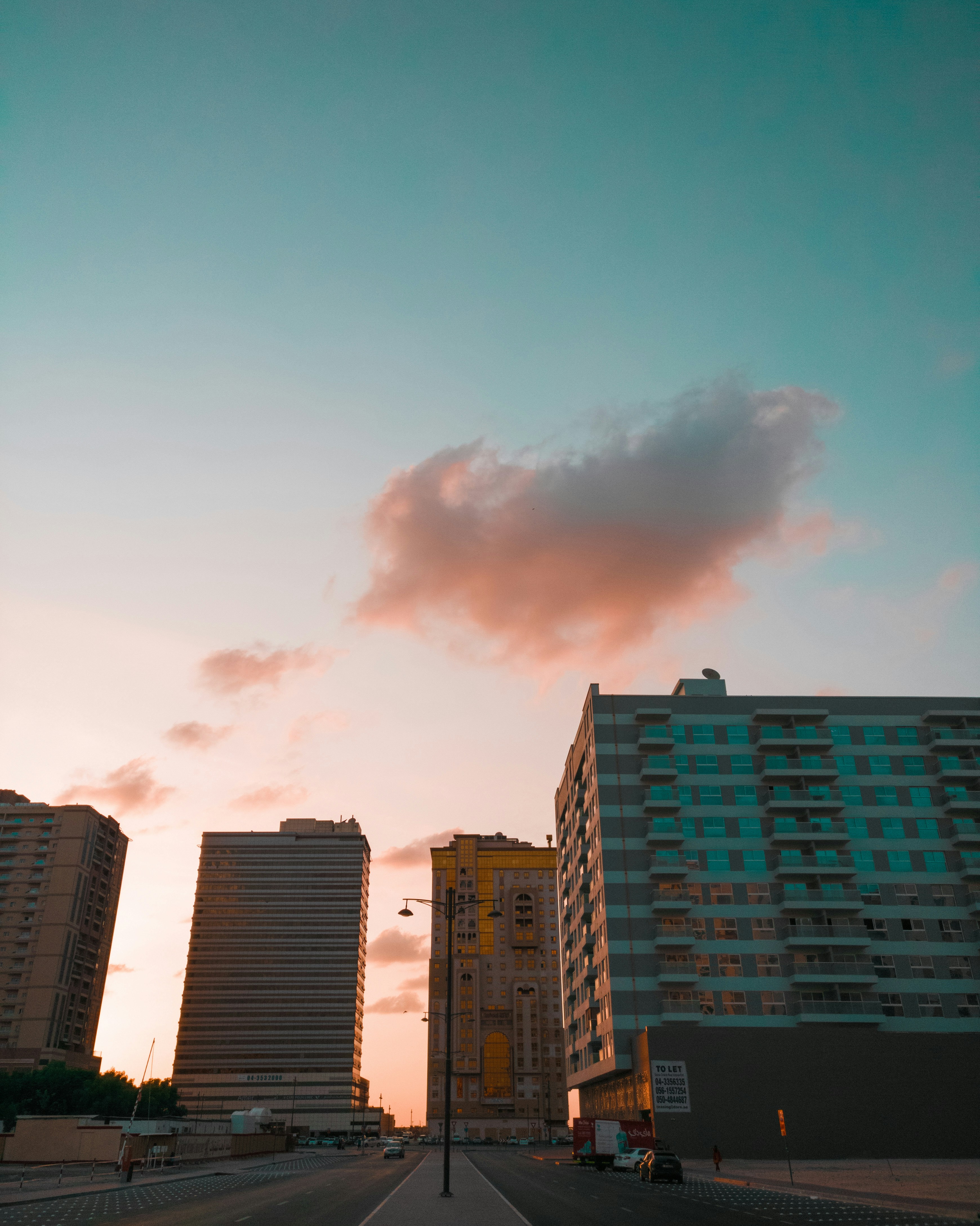 Sunset colors illuminate a city skyline dominated by turquoise and gray high-rises, with a calm street level below.