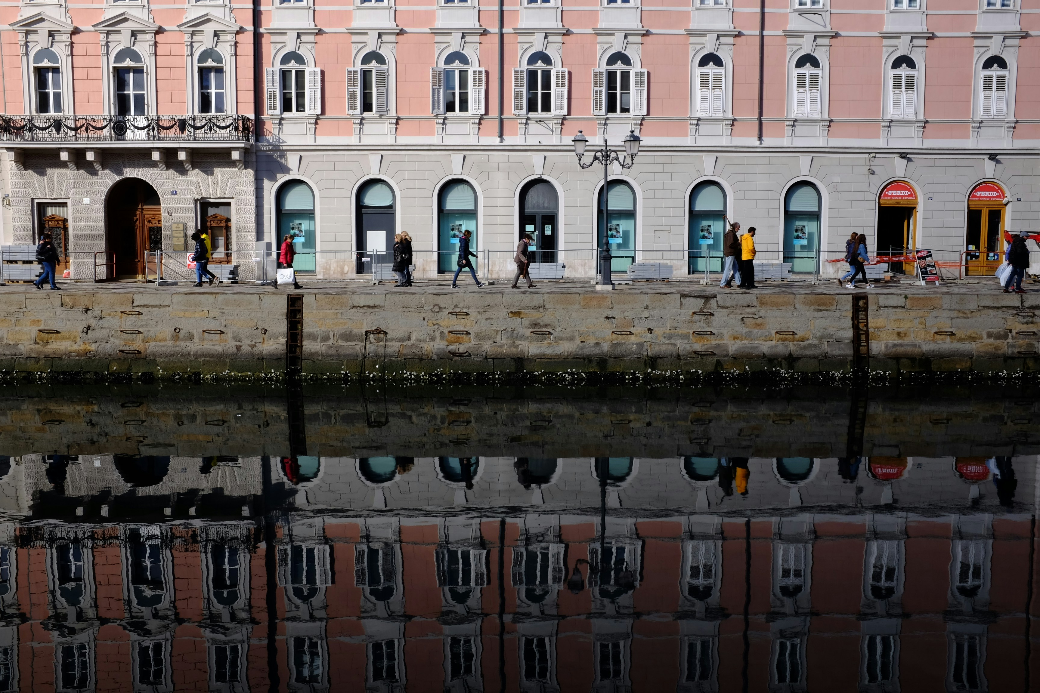 People strolling by a canal with a pink facade and window reflections in the water.