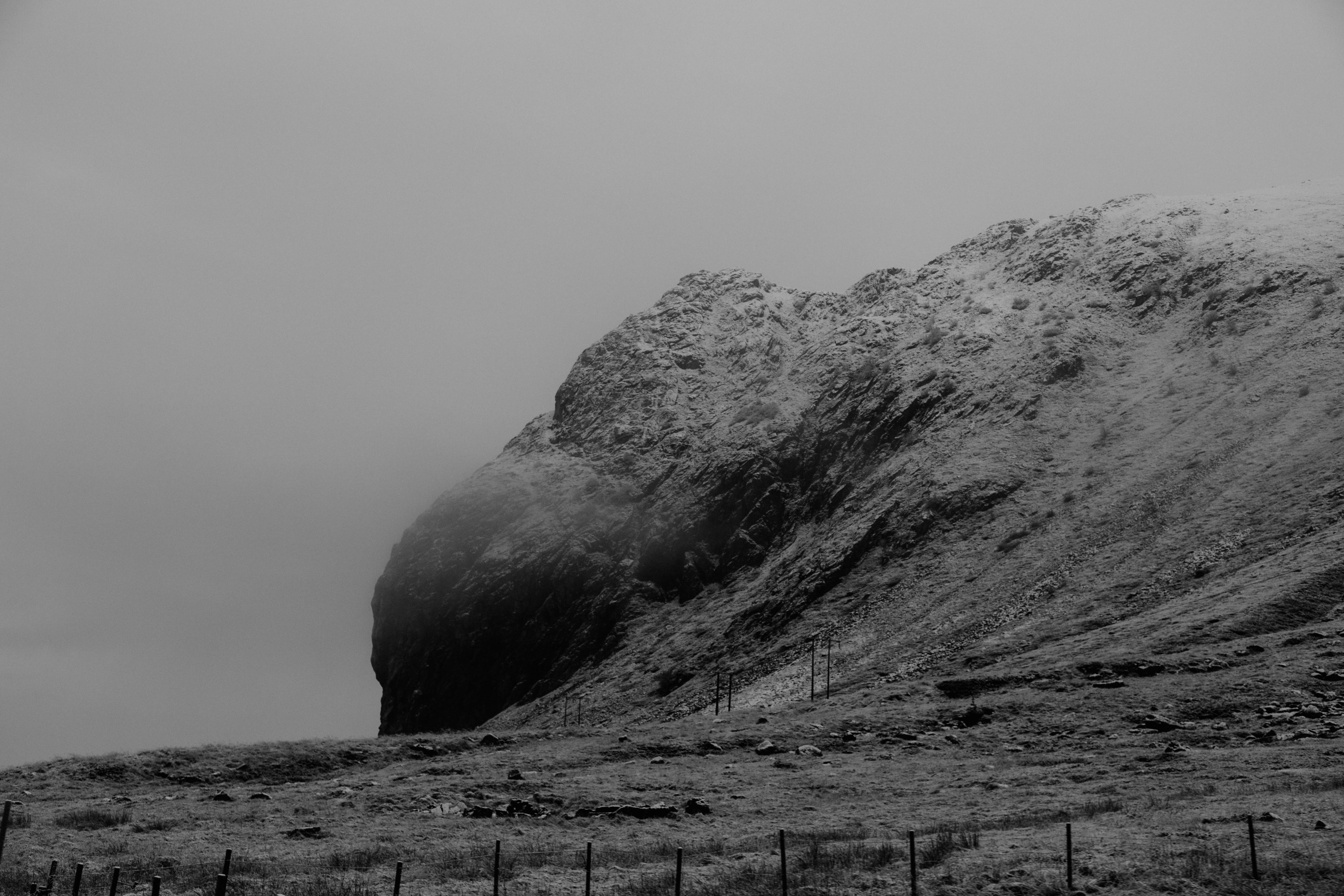 A rugged mountain shrouded in mist, showcasing the stark beauty of the landscape in monochrome. The foreground features a grassy plain with distant fence posts.