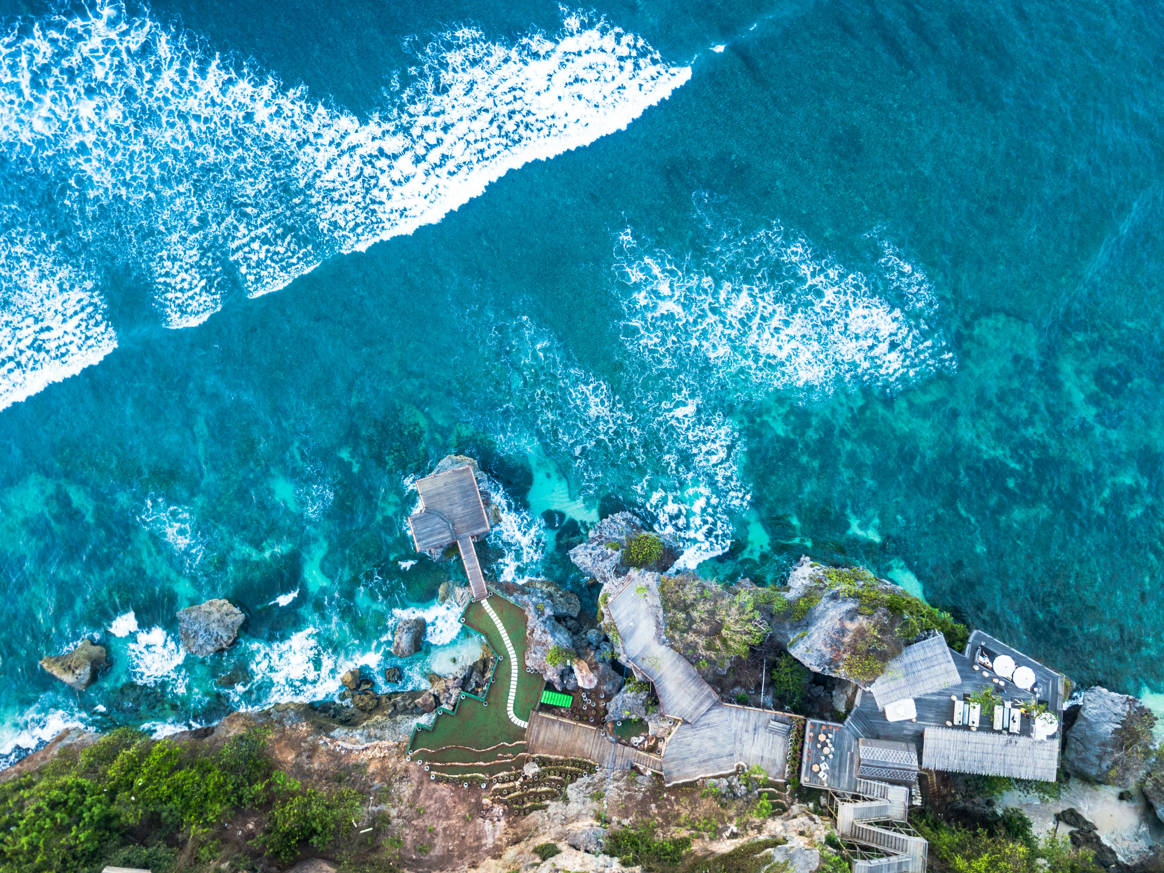 Aerial view of ocean waves meeting rocky coastline with scattered buildings on a clear day.