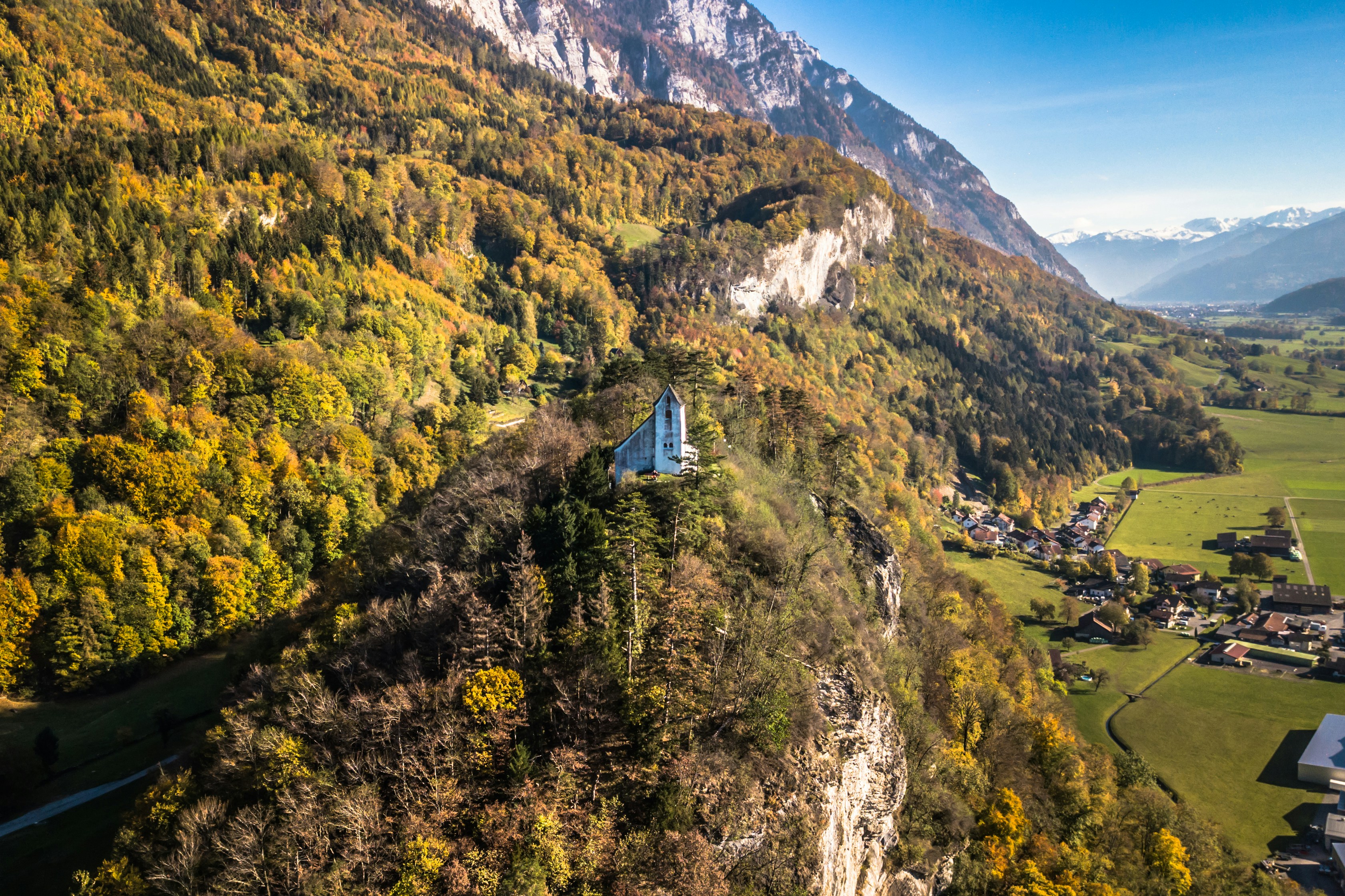 A quaint church perched atop a rocky outcrop surrounded by vibrant autumn foliage and expansive valleys. The scene captures the harmony between nature and architecture.