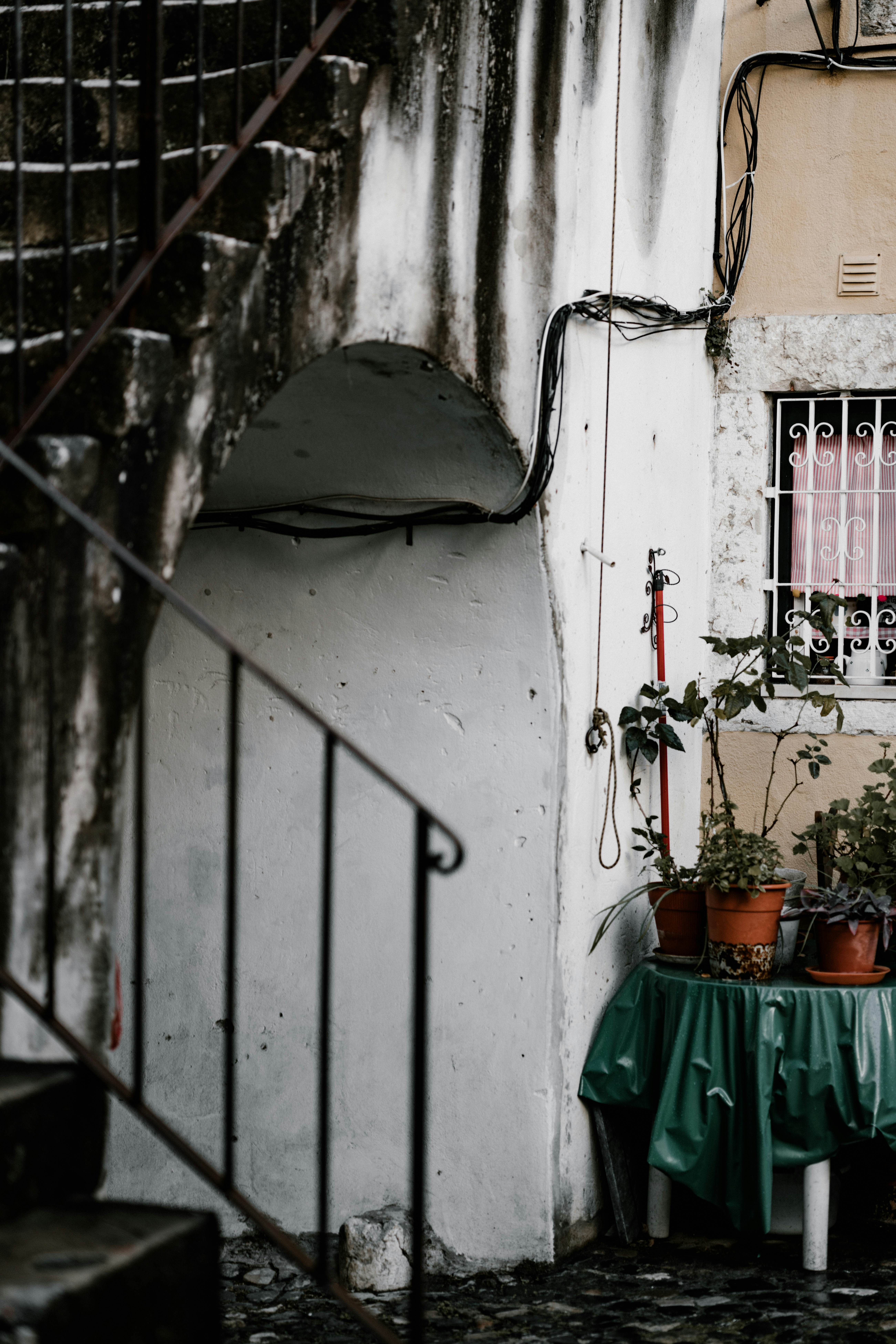 An inviting green table adorned with potted plants sits beneath a weathered archway, framed by a textured wall and a window with delicate ironwork.