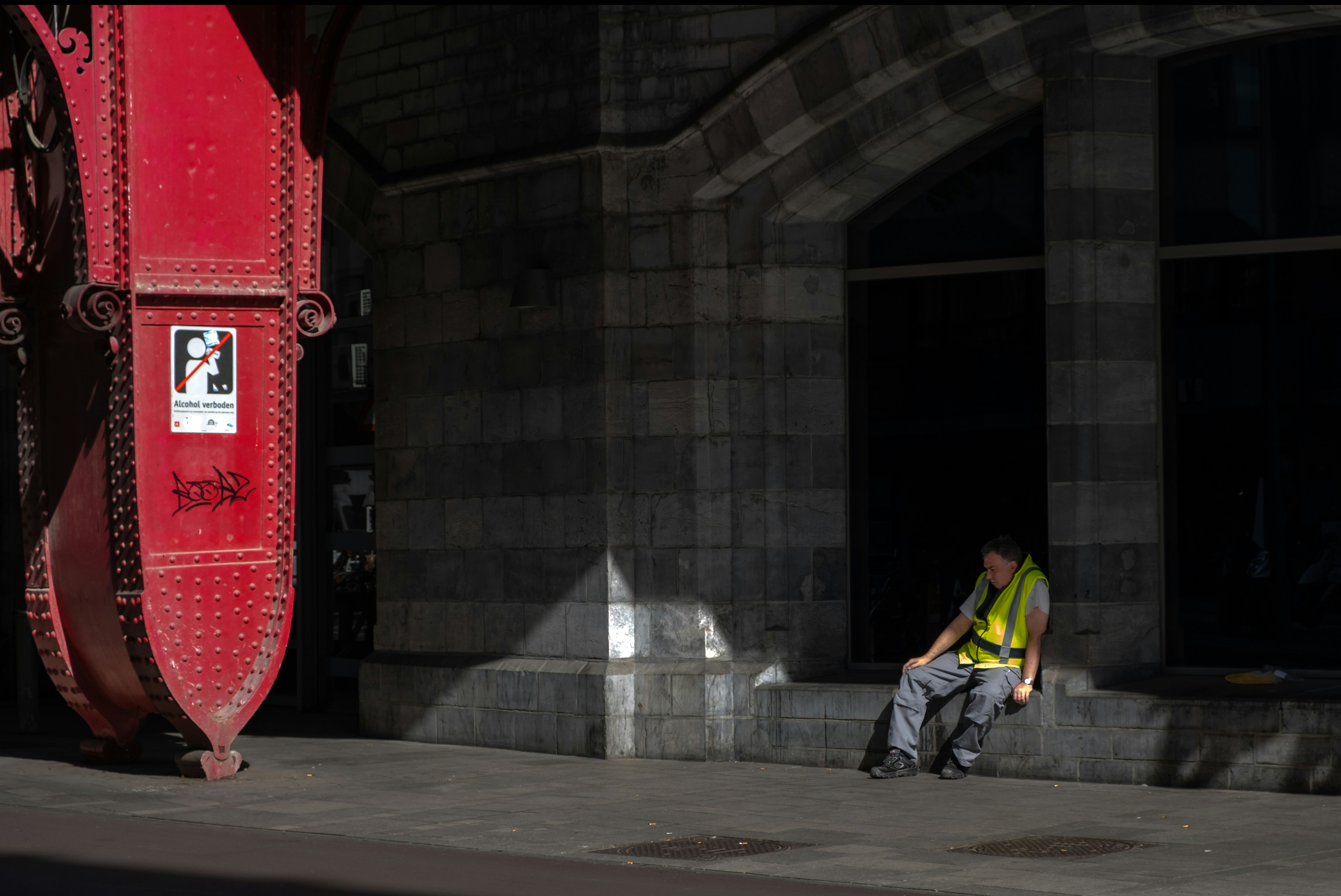Person resting on a bench in a shadowed urban area with bright red architectural details.