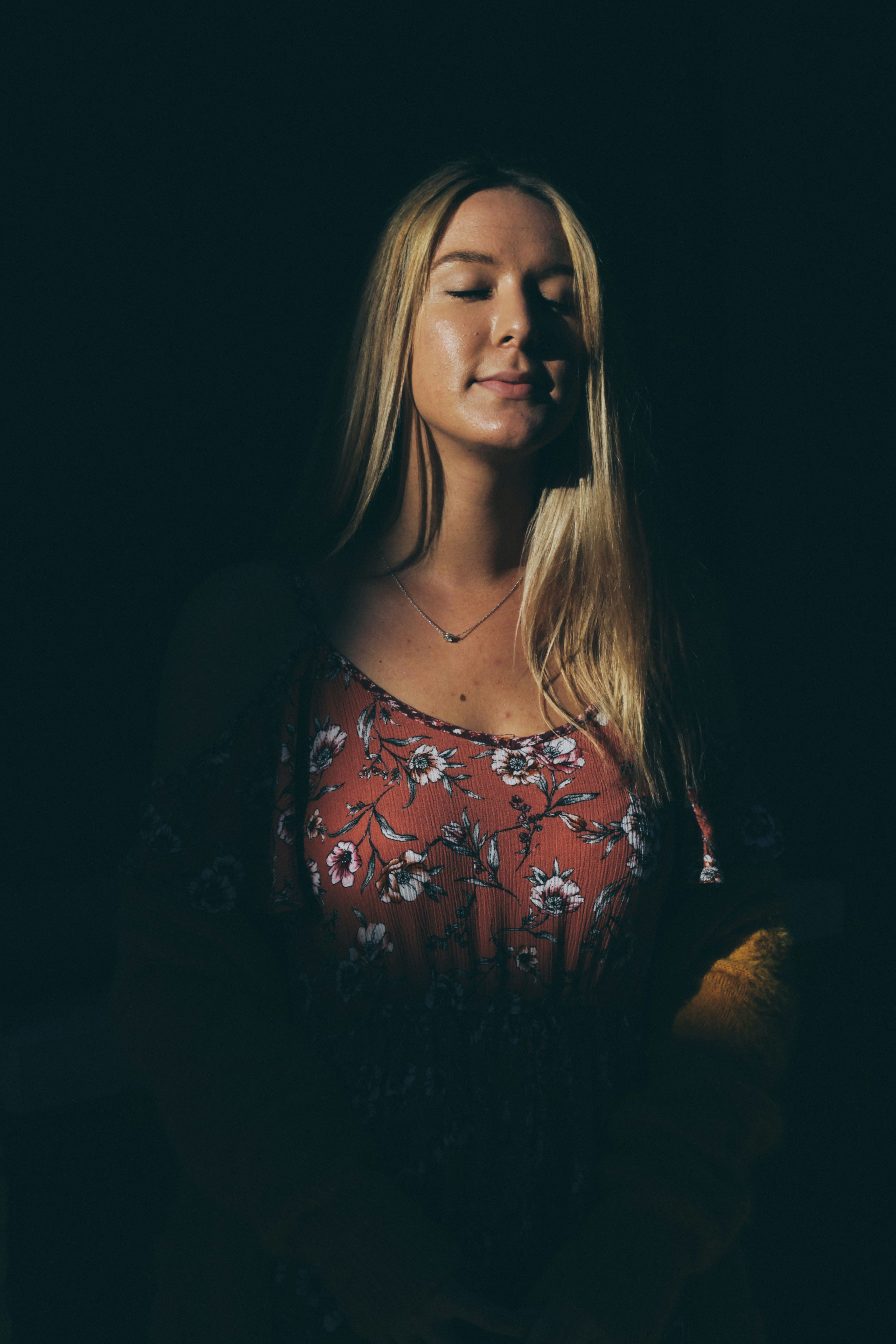 A woman with closed eyes stands in soft light, her floral dress contrasting against a dark background. The interplay of light and shadow creates a tranquil atmosphere.