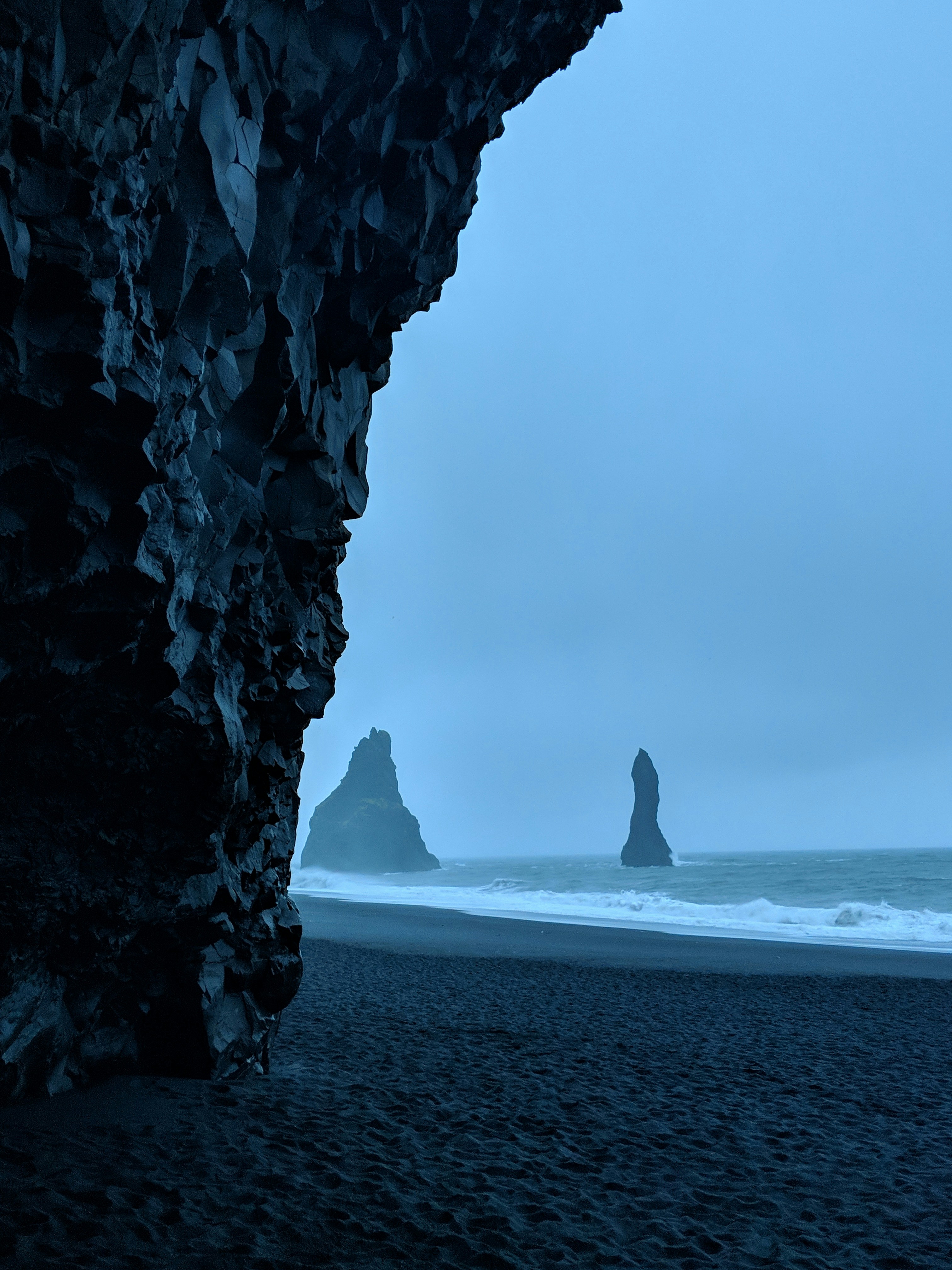 Jagged black rock formations tower over a misty beach with sea stacks rising from the ocean under a moody sky.