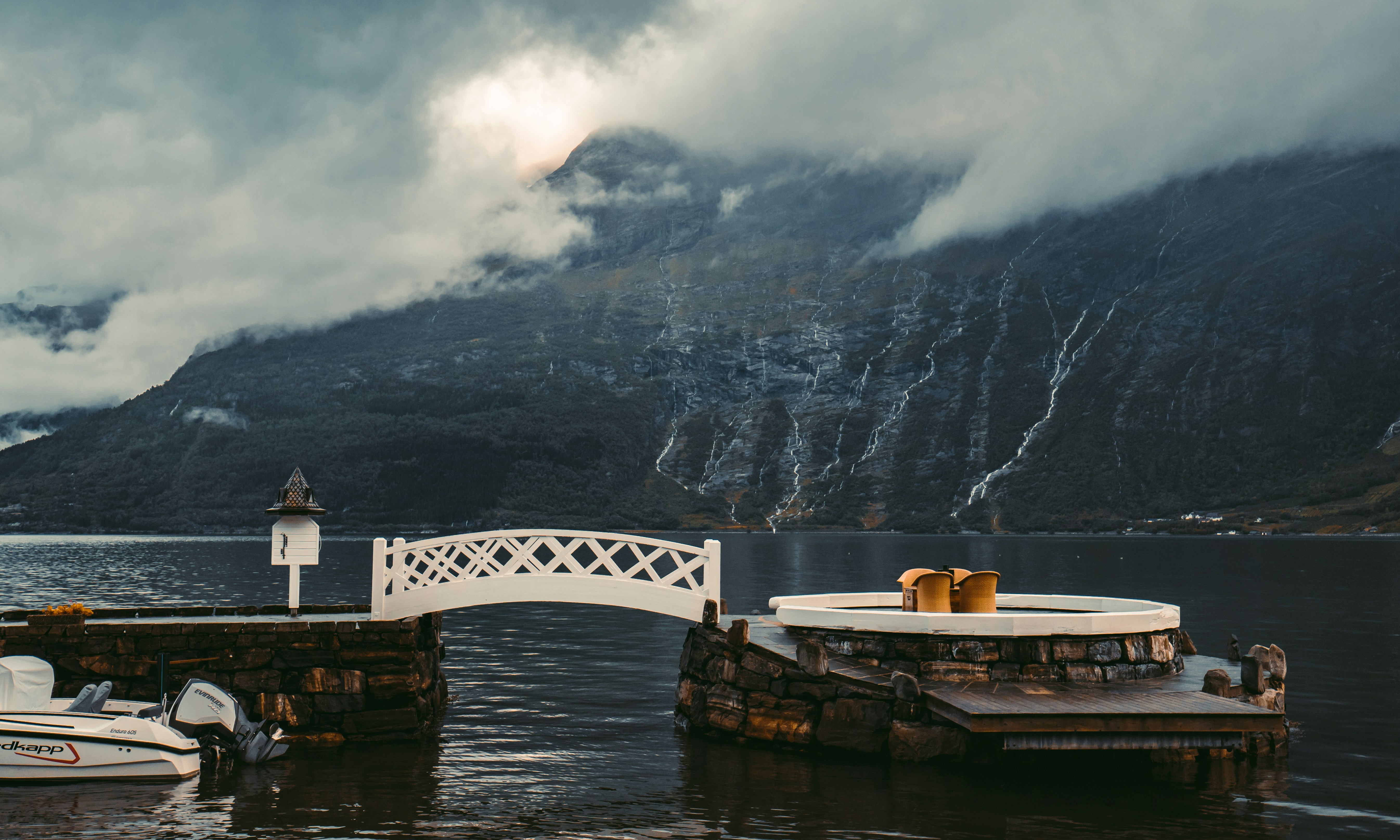 Wooden dock with a small boat and bridge set against misty mountains and a tranquil lake.