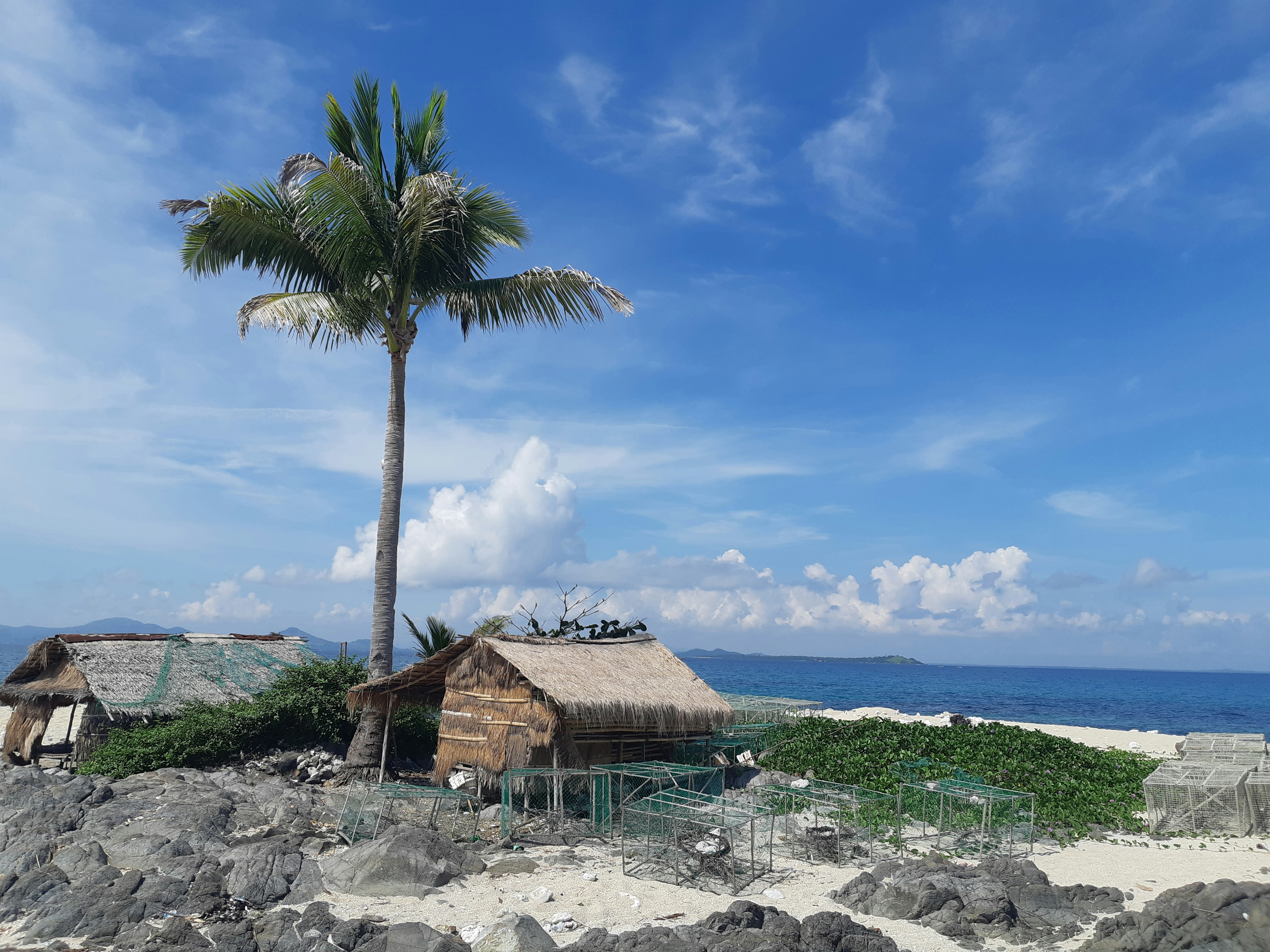 Tall palm and thatched huts frame a rocky beach beneath a bright blue sky, with the calm sea stretching to the horizon.