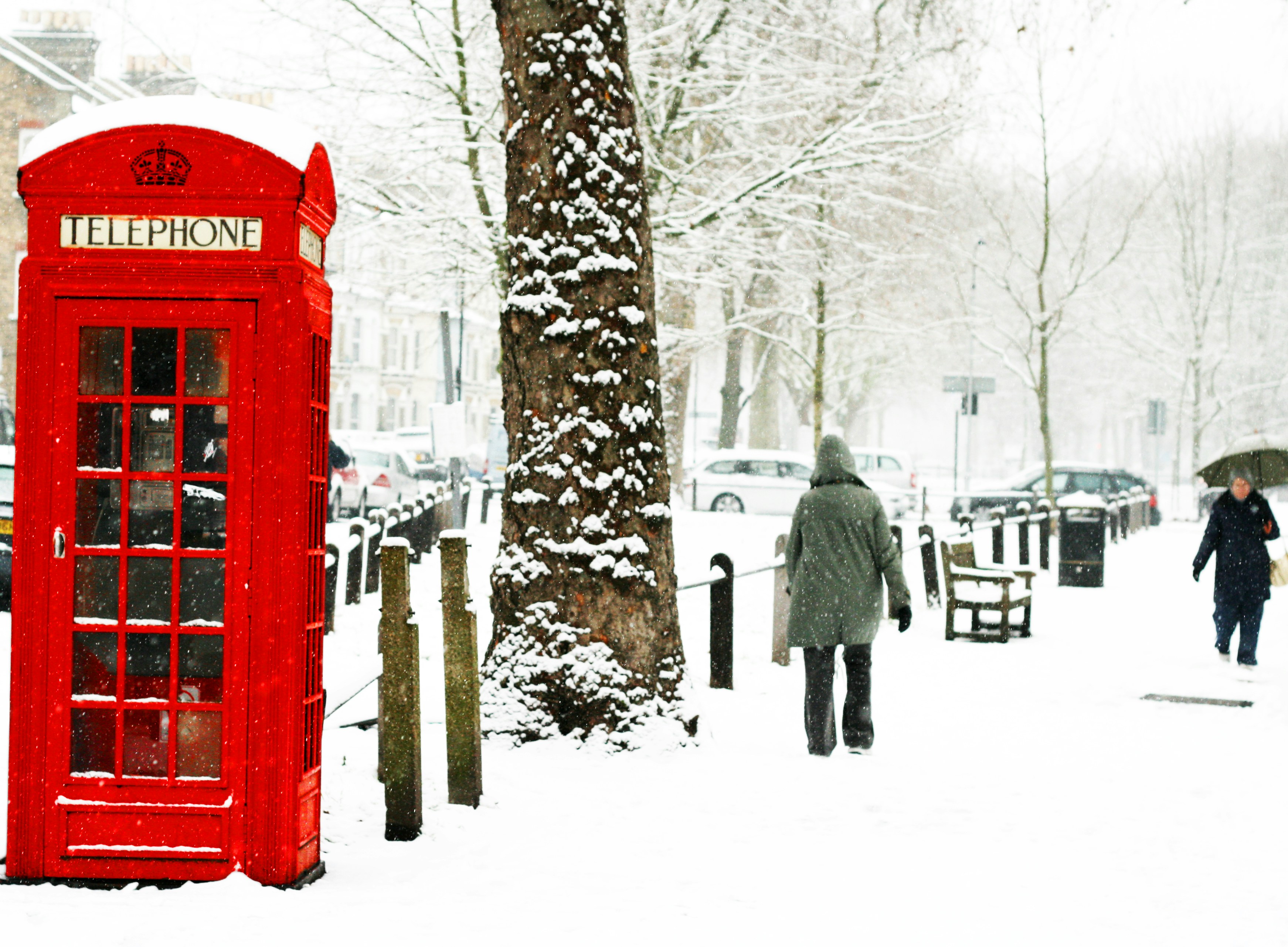 Red phone box stands out against a snowy London street, flanked by snow-dusted trees and passersby.