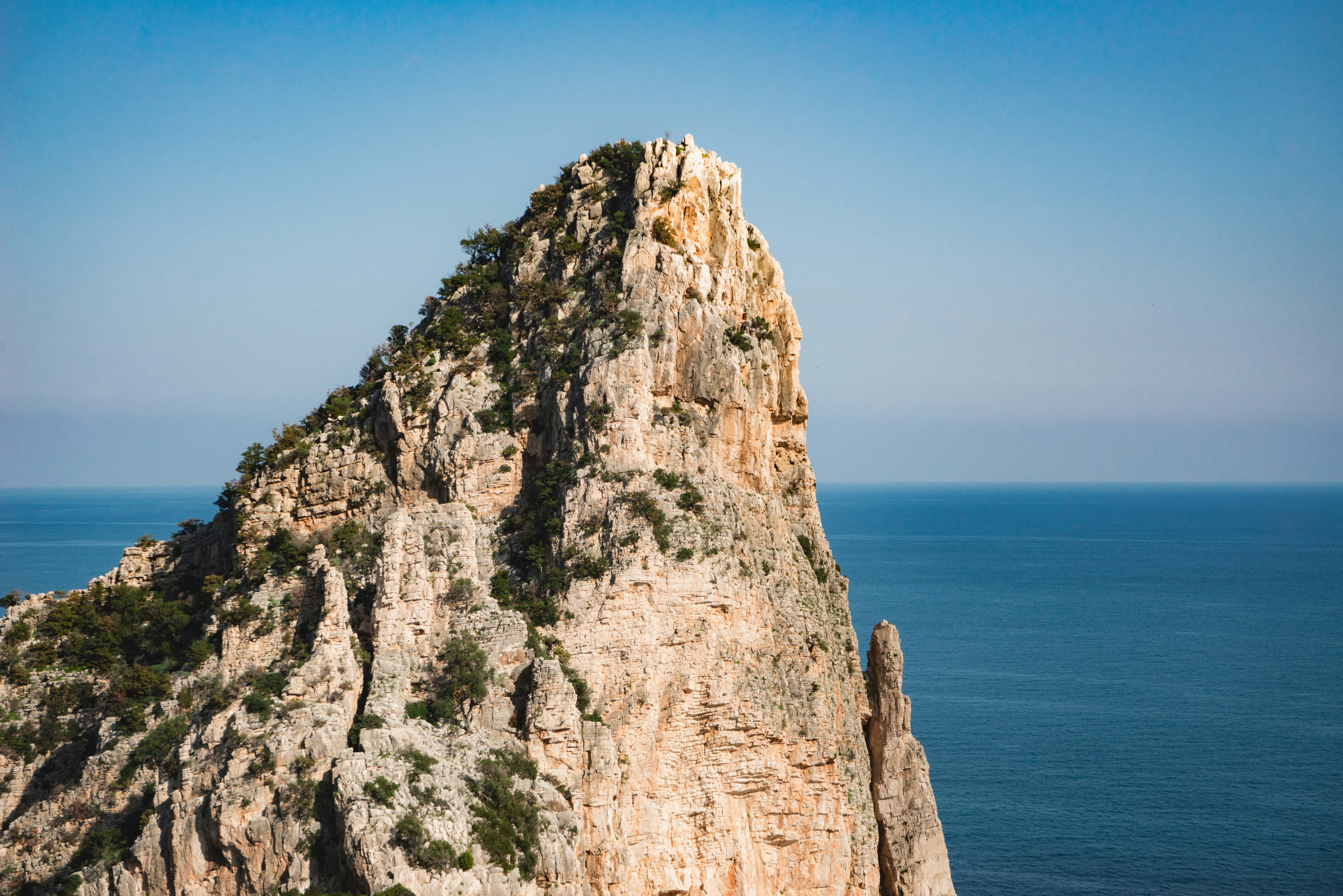 Prominent rock formation rising sharply against a clear blue sky and tranquil ocean backdrop.