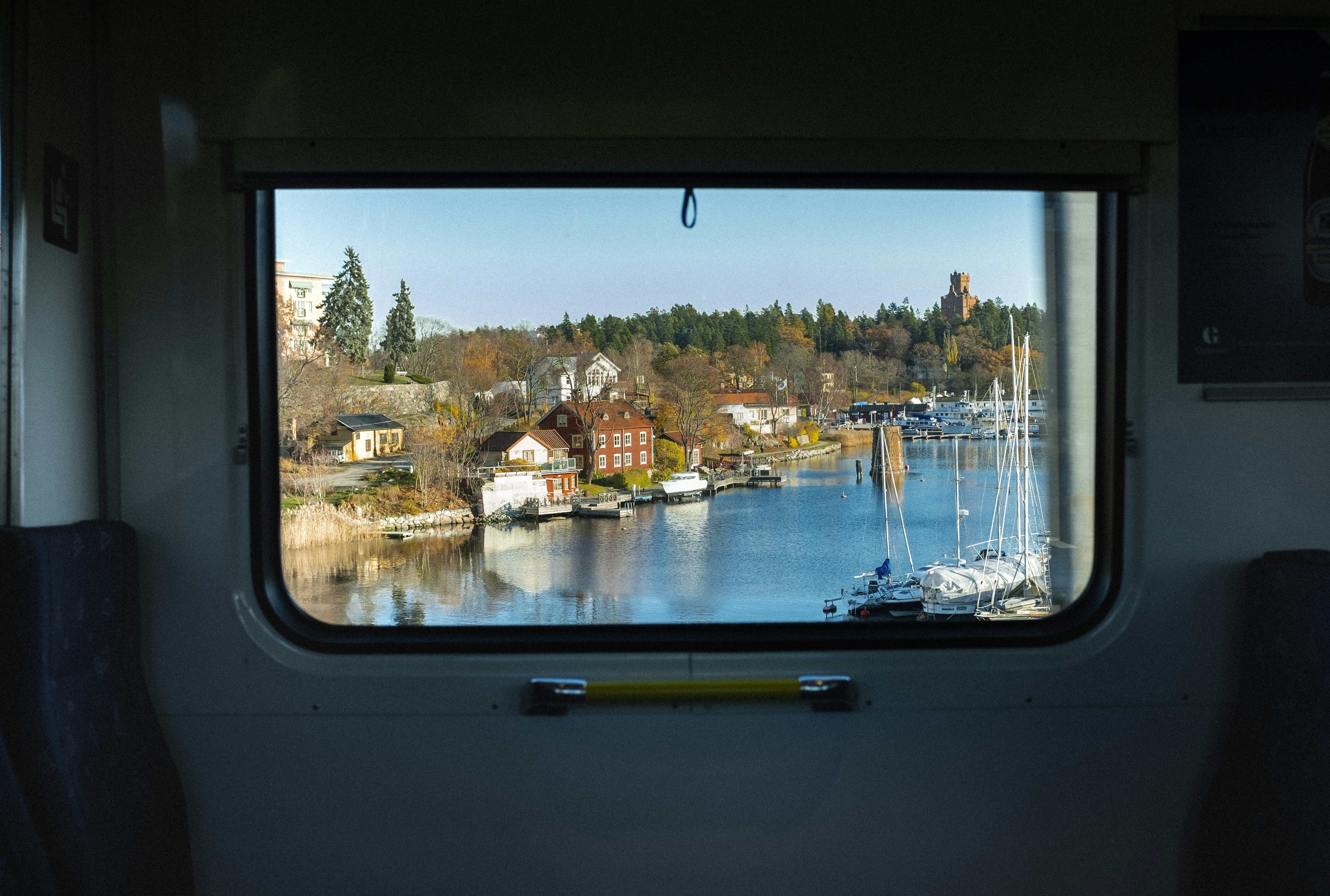 A tranquil waterfront scene framed by a train window, showcasing charming houses and moored boats along a serene riverbank.