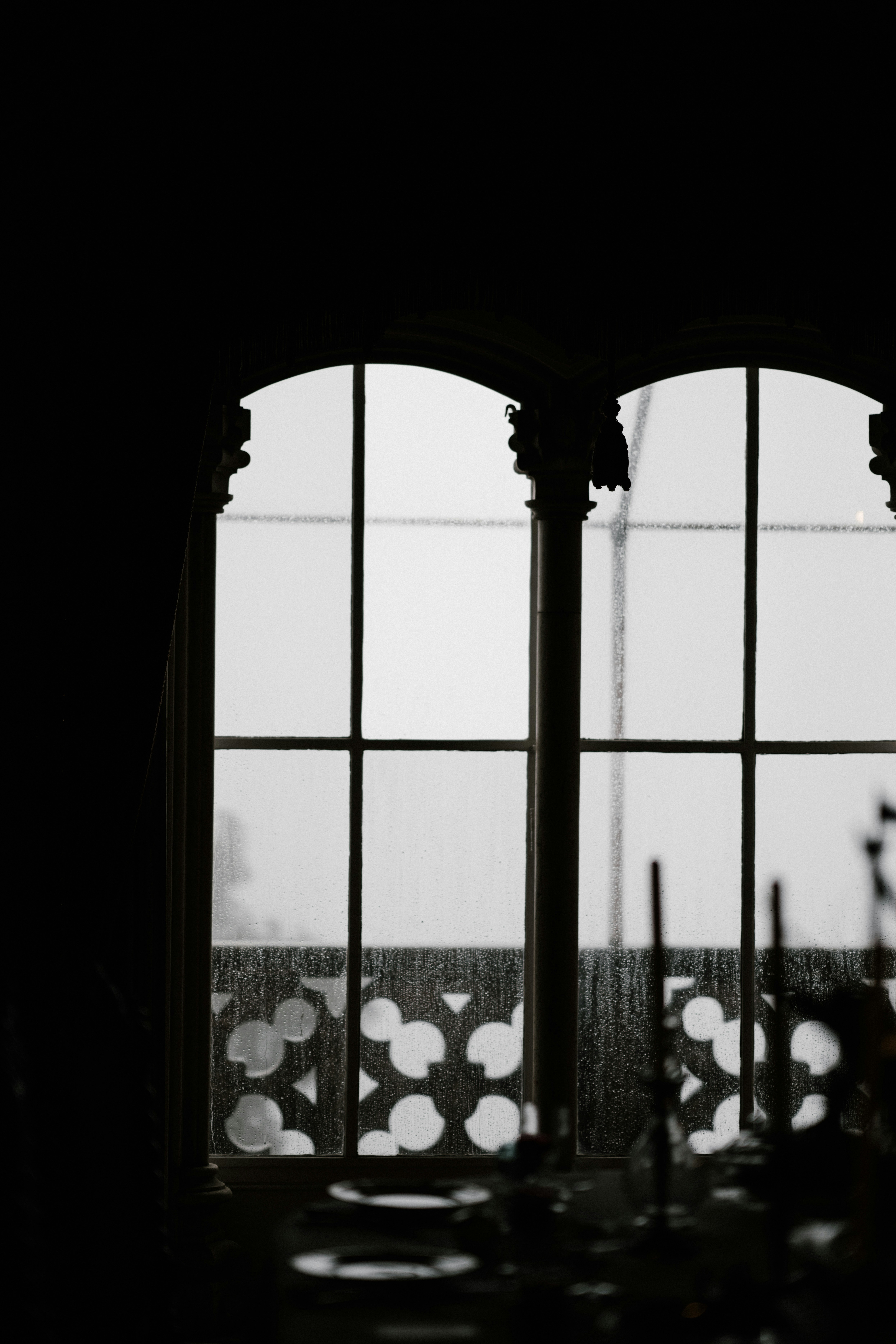 Raindrops cling to a windowpane, obscuring a view of a patterned balcony beyond. The interplay of light and shadow creates a moody ambiance.