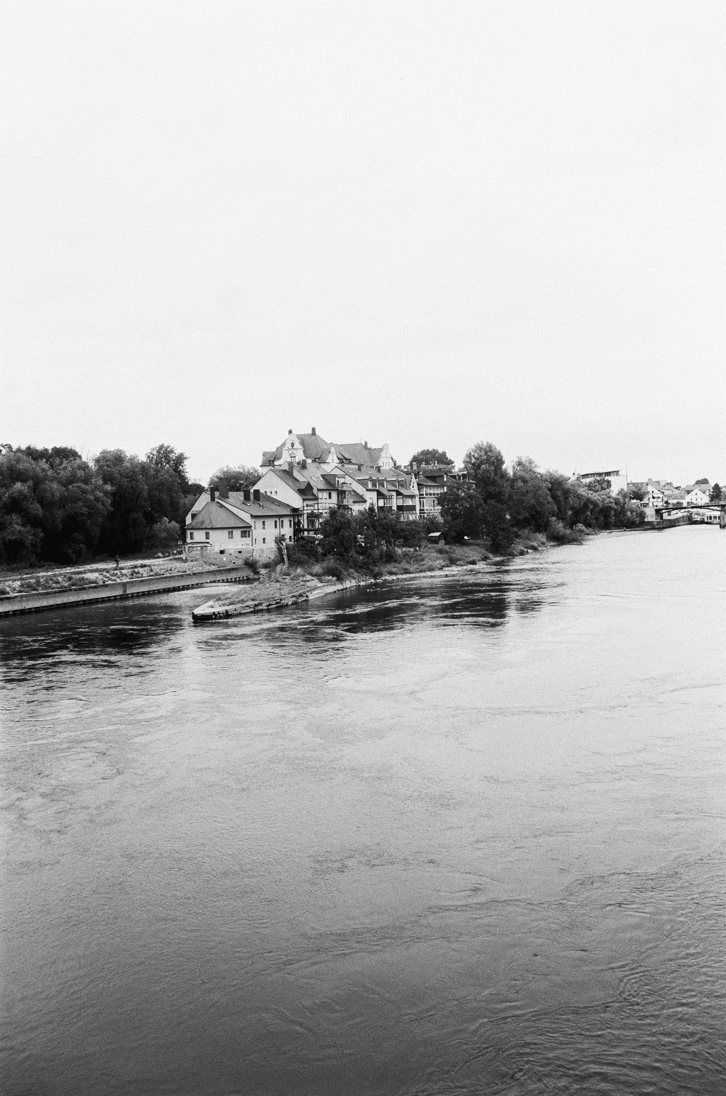 Monochrome photograph of a riverside village clustered along a gentle bend, with calm water and leafy banks.
