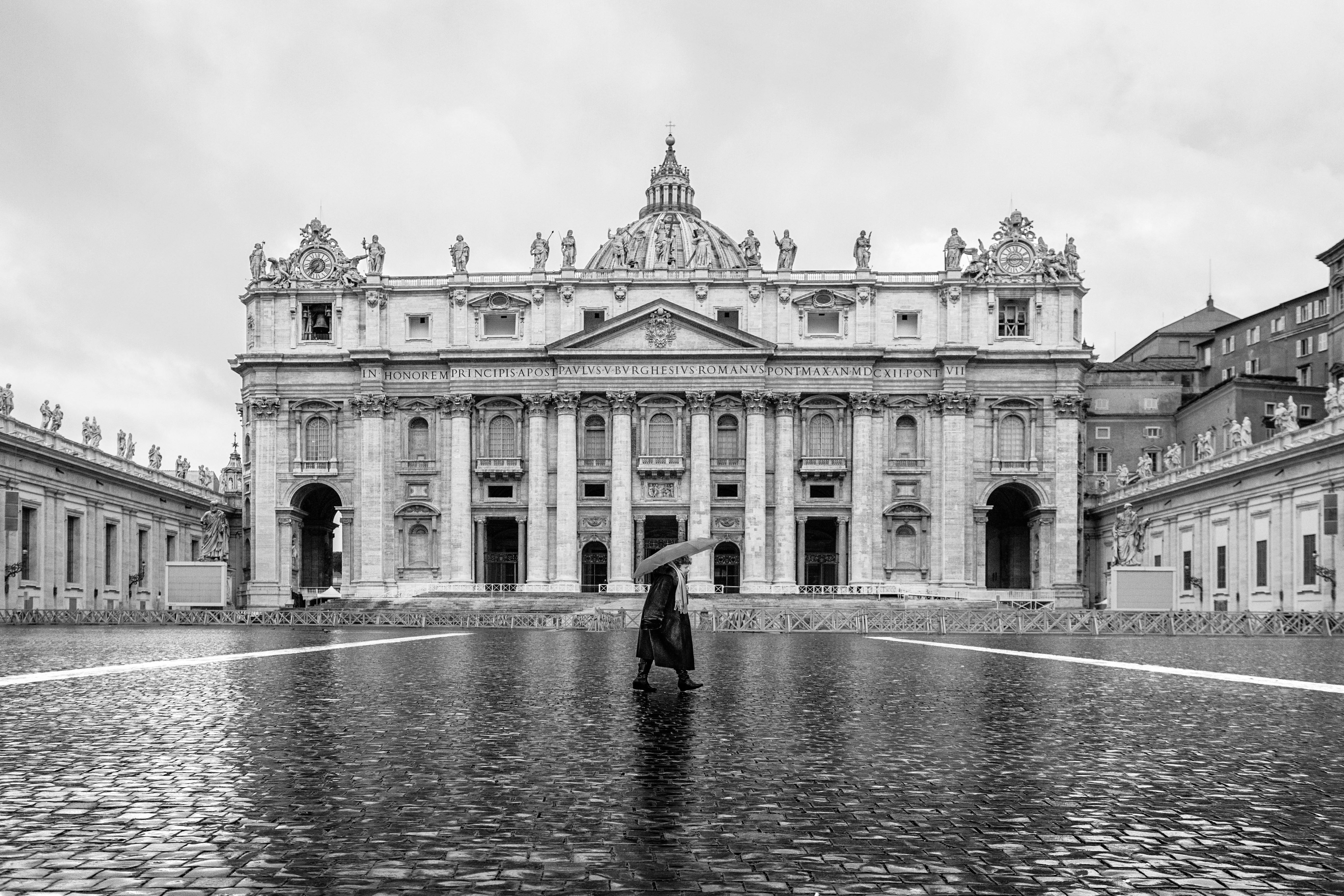 Individual walking with an umbrella in front of the grand St. Peter's Basilica, captured in black and white.