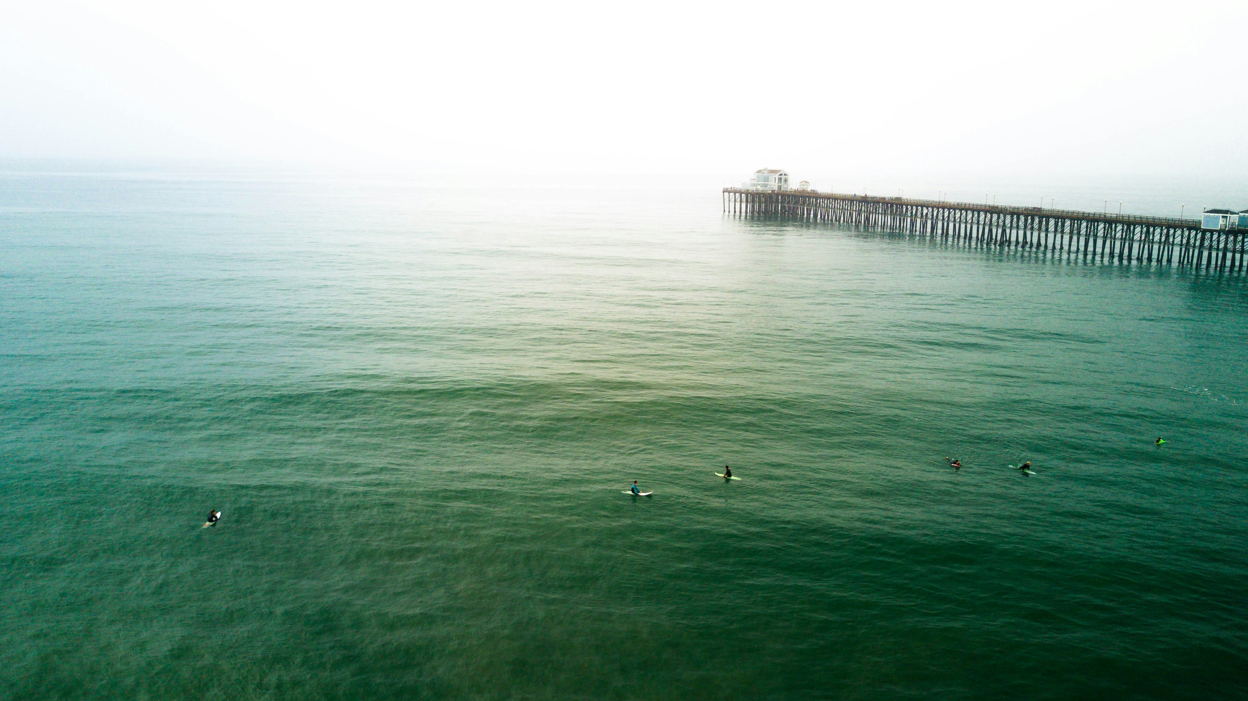 Surfers floating in calm ocean waters near a distant pier under a soft, overcast sky.