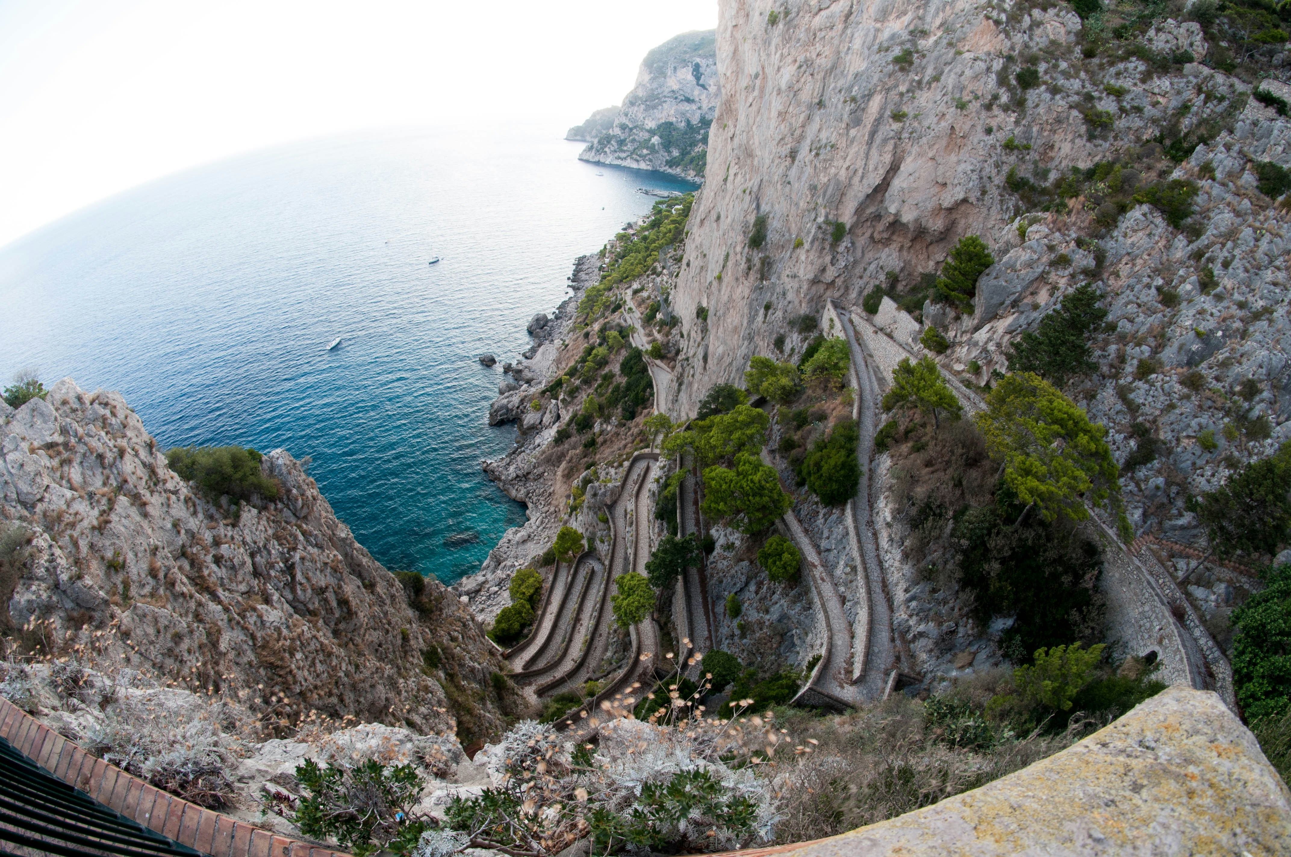 Aerial view of a serpentine pathway leading down a rocky cliffside to the tranquil sea below, framed by lush greenery.