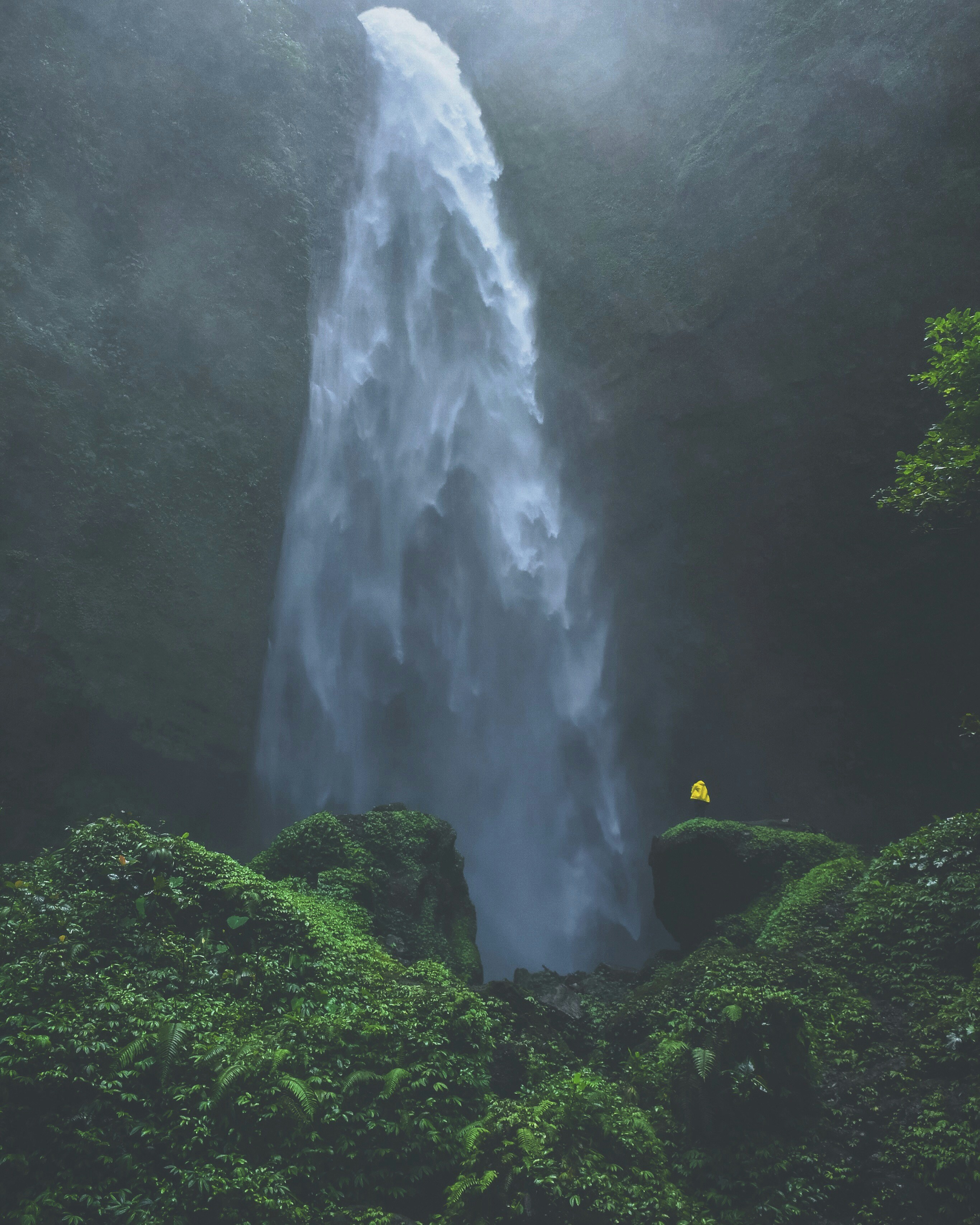 Tall waterfall plunging into a verdant canyon, with mist rising and a person in a yellow jacket for scale.