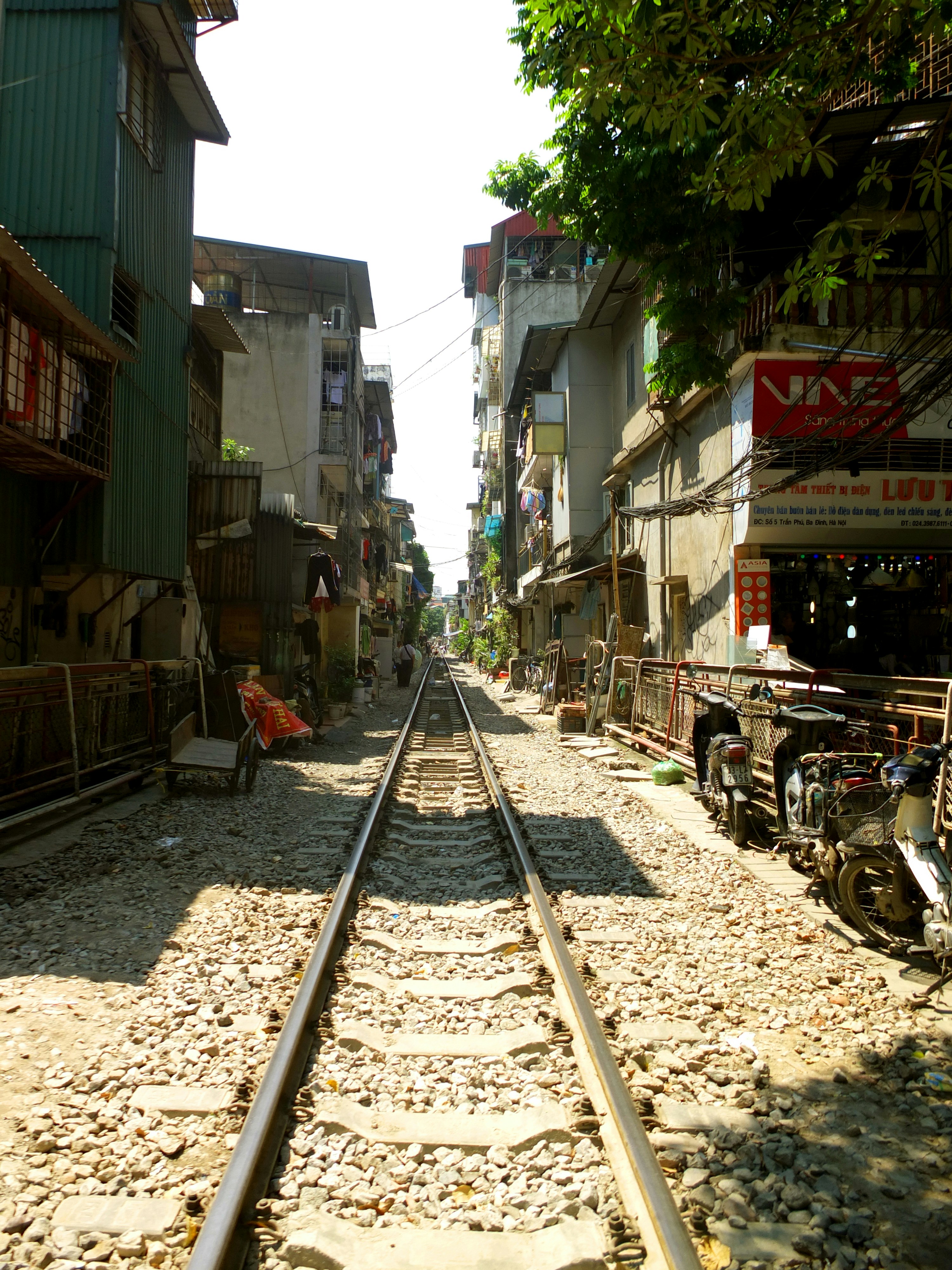 Narrow railway tracks stretch down a bustling urban alley flanked by buildings and shops. The scene captures the essence of city life intertwined with transportation.