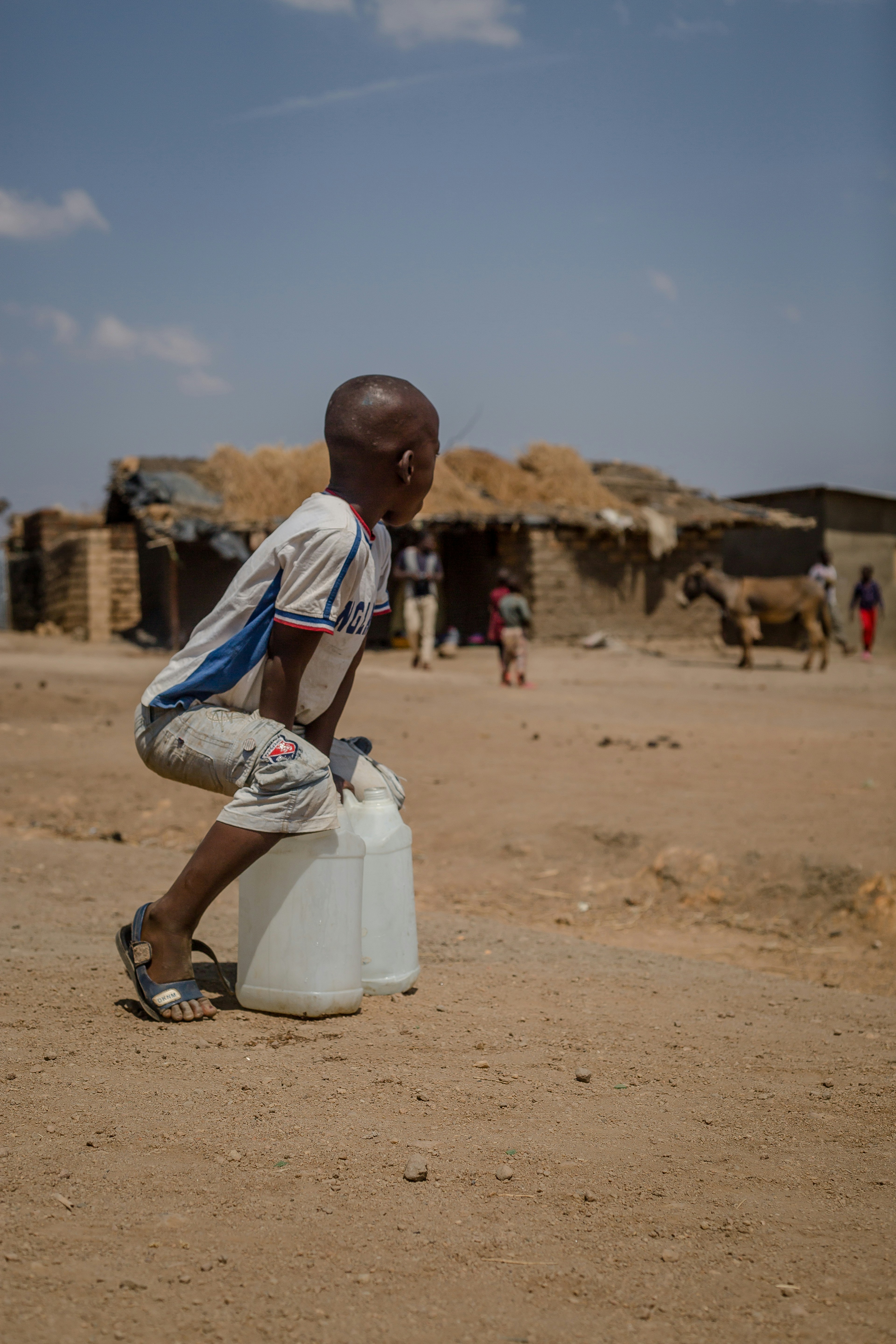 A young boy balancing two water containers on his knees in a rural setting, with rustic buildings and a few people in the background. The scene captures the daily struggle for water in a community.