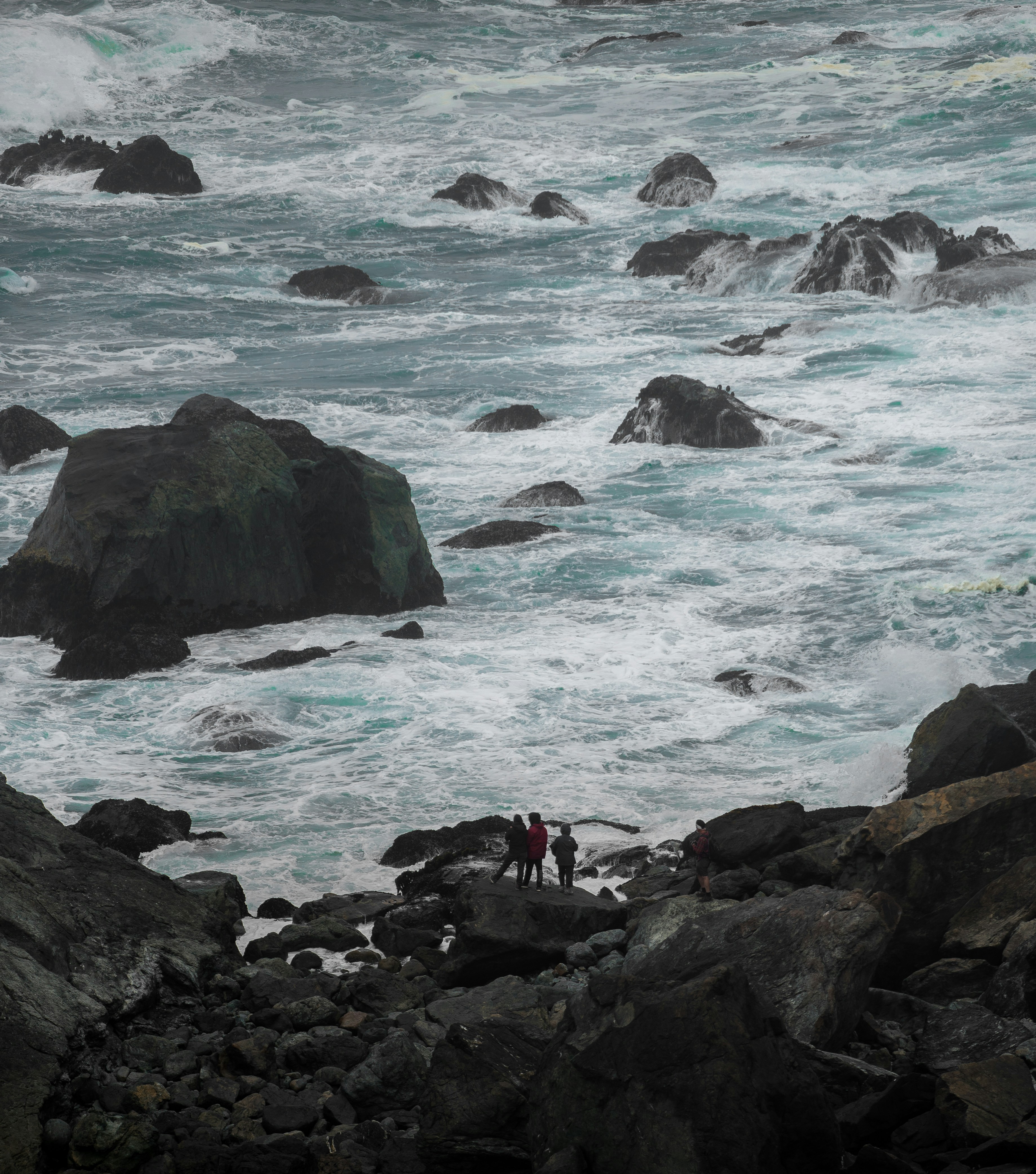 Two figures standing on rocky shore as waves crash around them, highlighting the power of the ocean. The scene captures a moment of human interaction with the wild landscape.