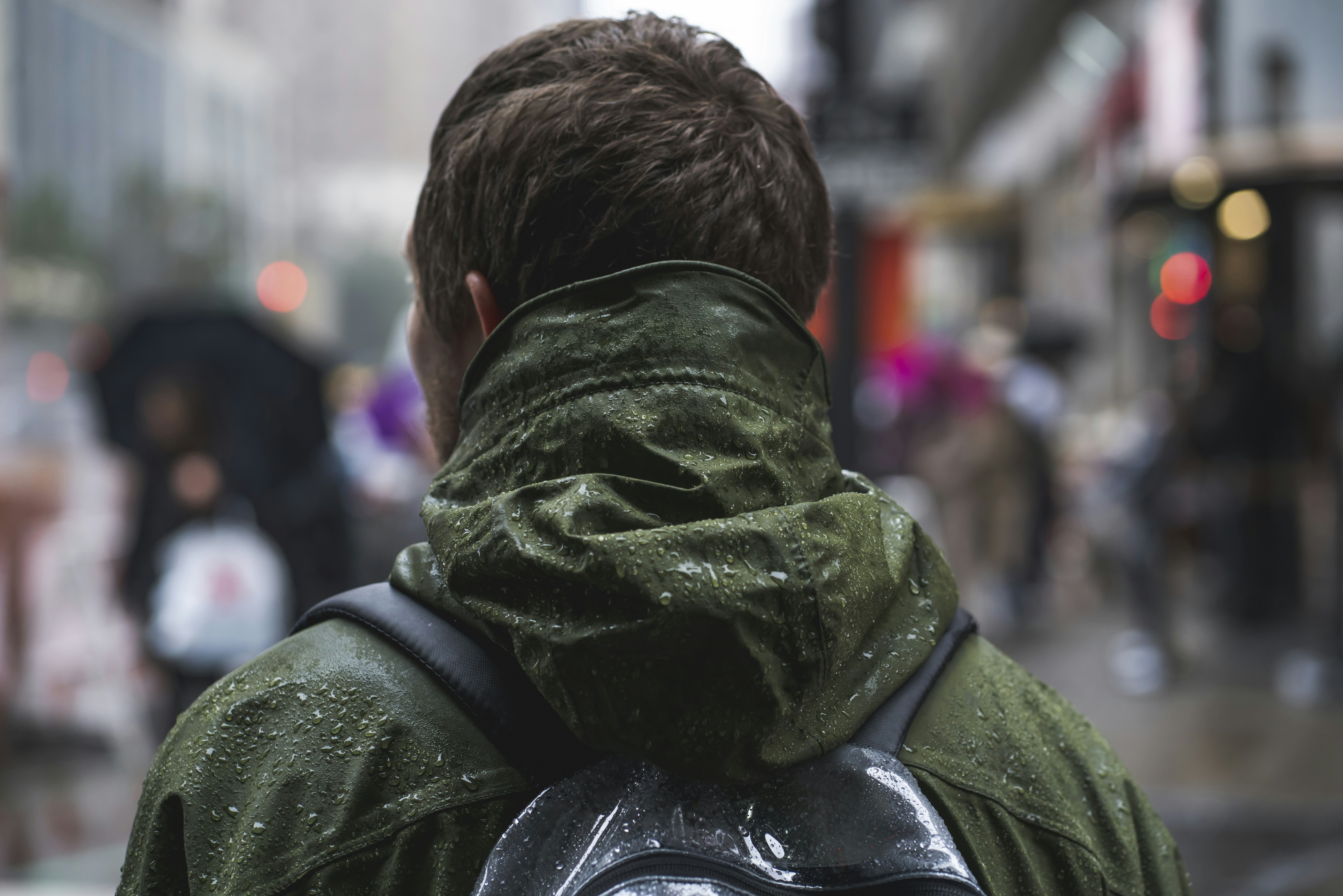 A figure in a rain-soaked green jacket stands amidst a bustling city street, with blurred umbrellas and lights creating a dynamic backdrop.