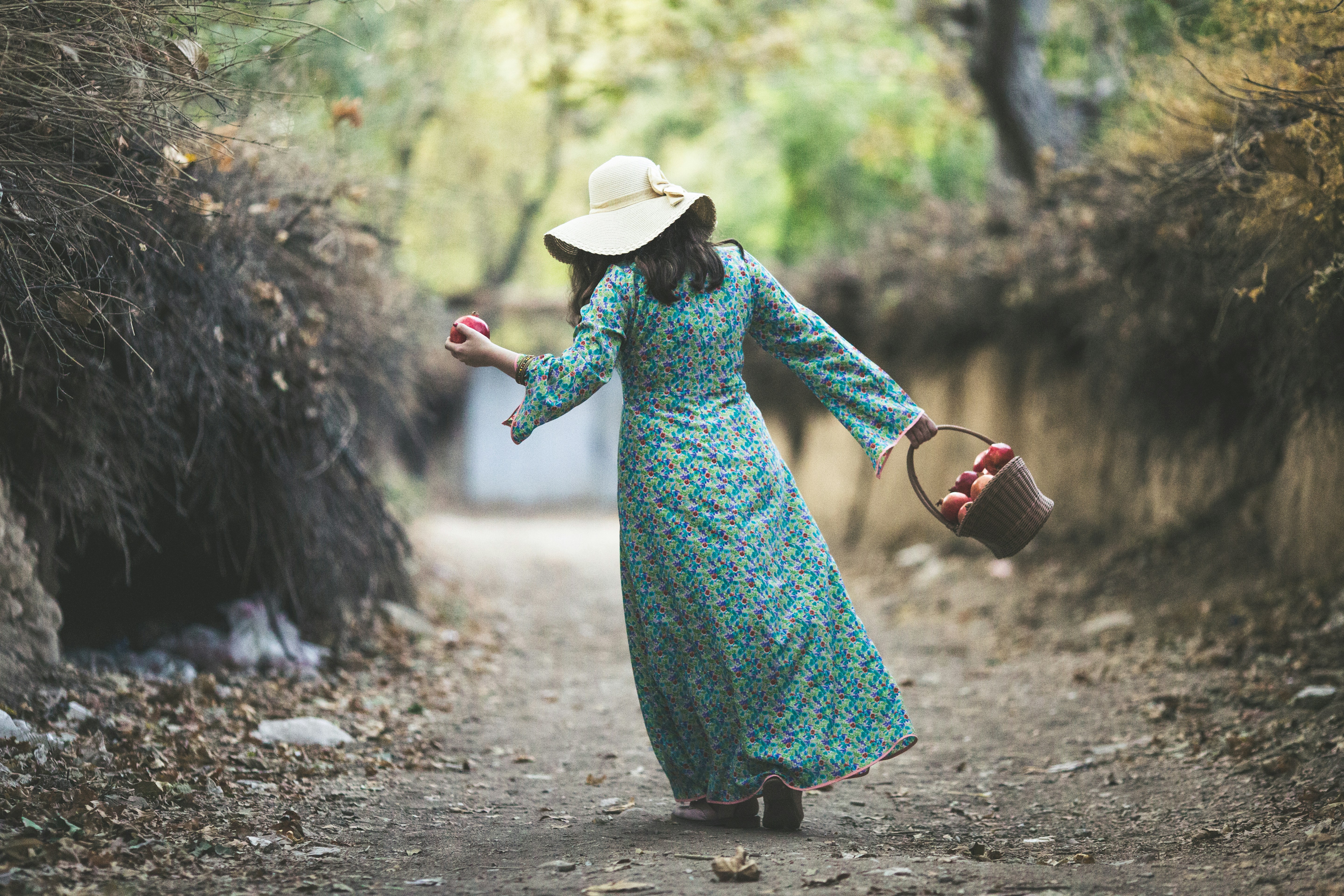 Woman in a flowing dress and wide-brimmed hat walks down a leaf-strewn path, holding a basket of apples.