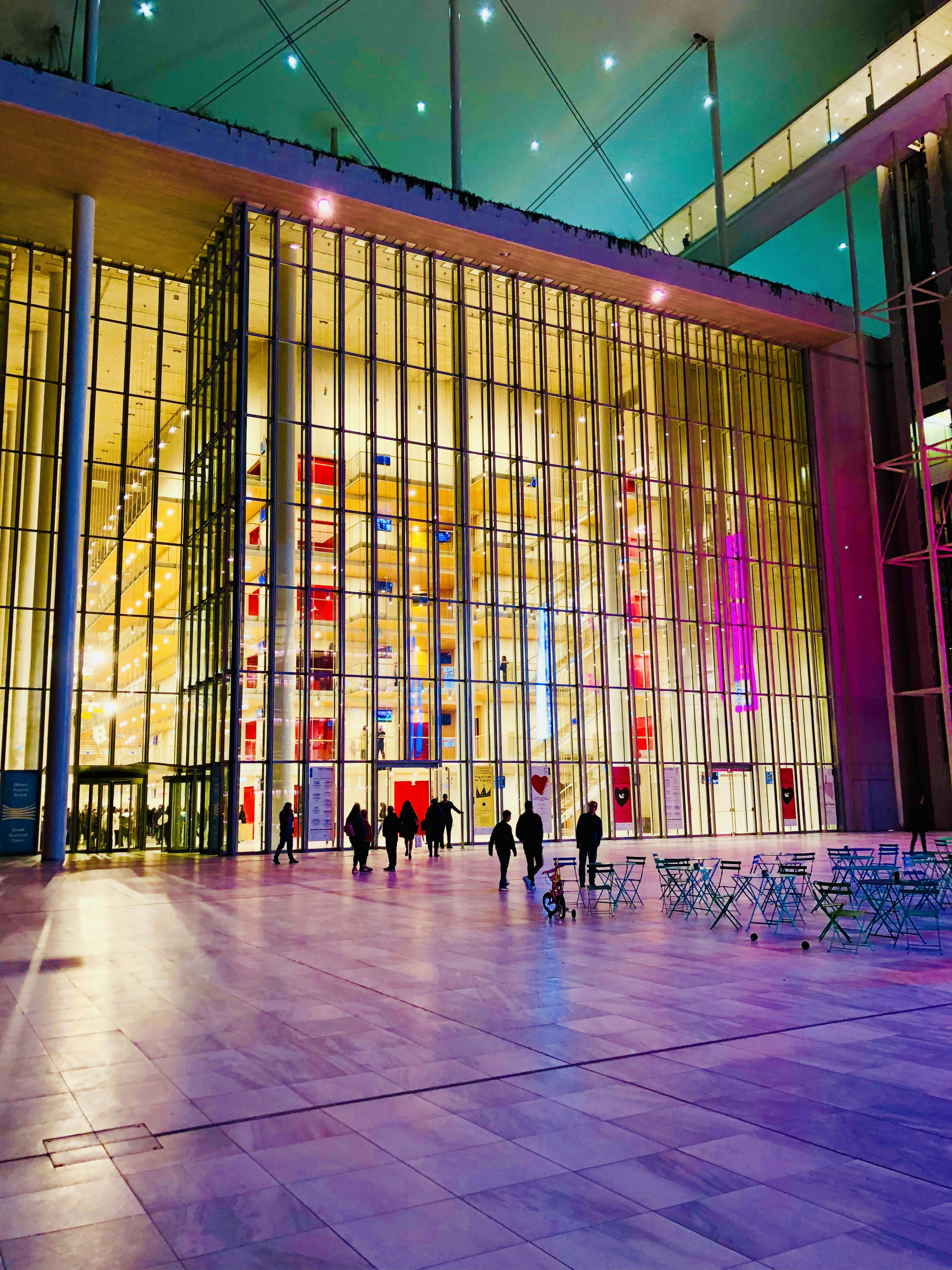 Vibrant glass facade of a modern building illuminated at night, showcasing colorful interior elements and silhouettes of people outside.