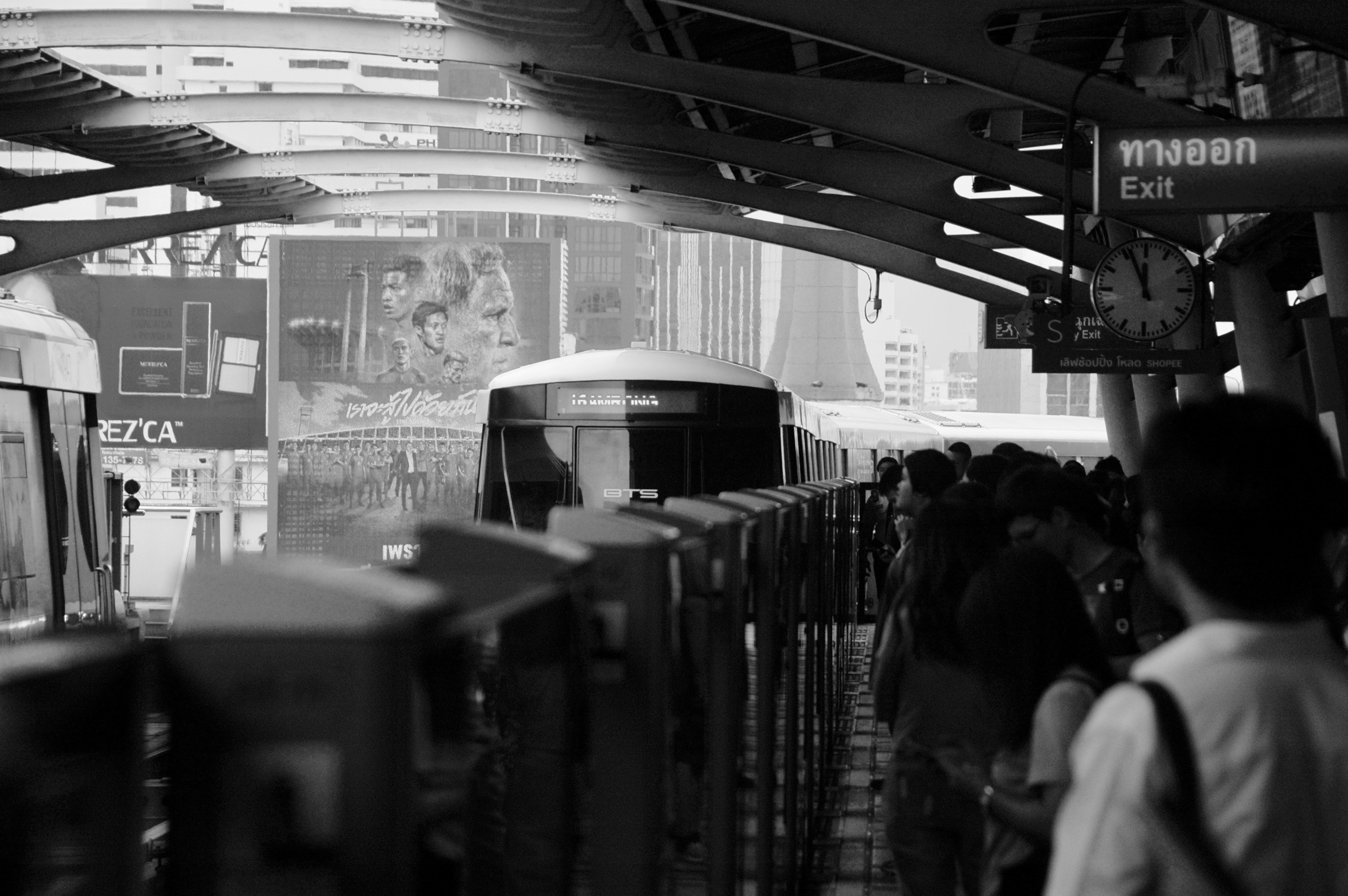 Crowd of commuters waiting at a bustling train station, framed by modern architecture and a large advertisement in the background.