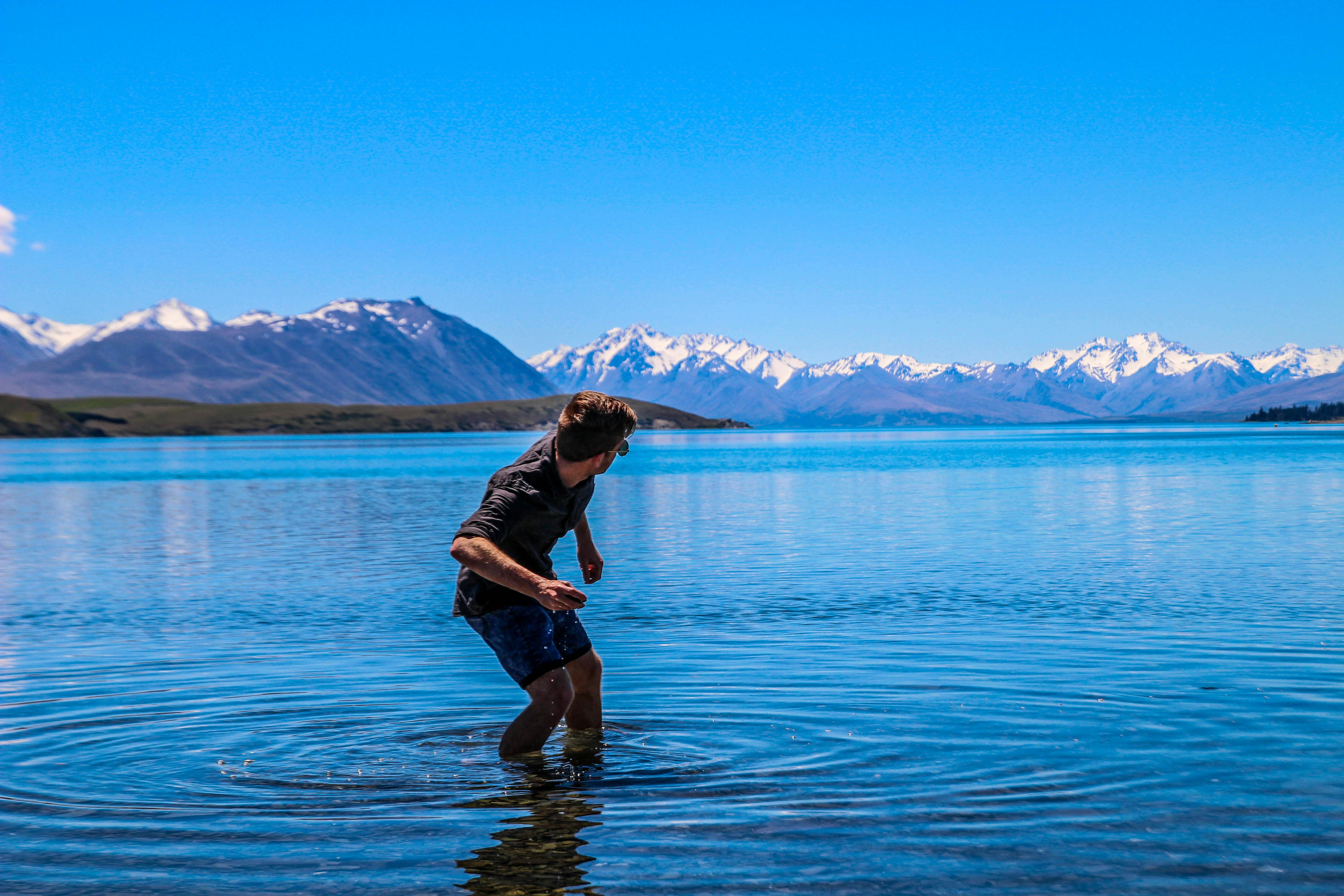 Person standing in a serene lake with snow-capped mountains in the background under a clear blue sky.