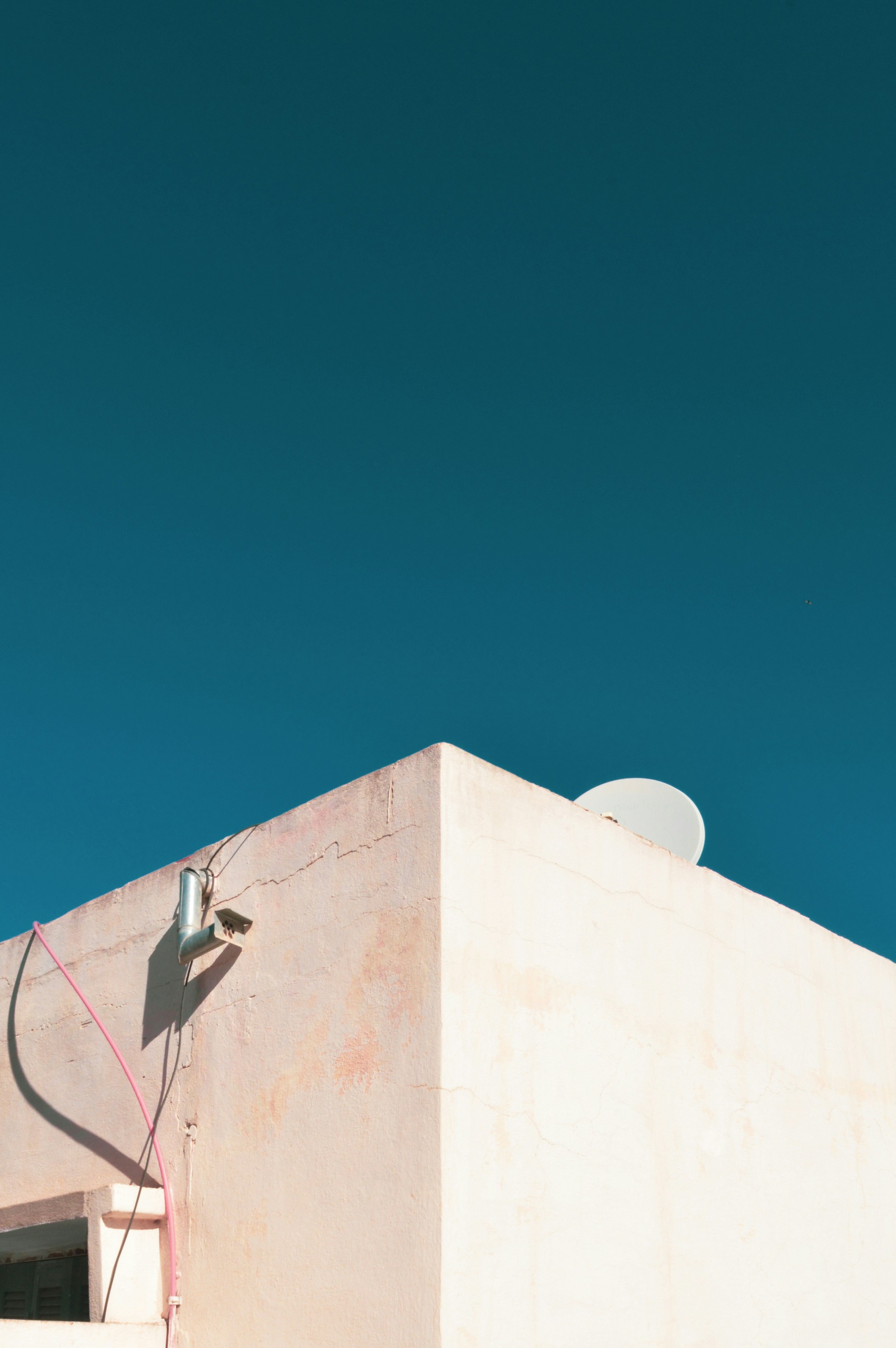 A corner of a white building against a clear blue sky, featuring a satellite dish and a pink cable. The simplicity highlights the interplay of color and form.