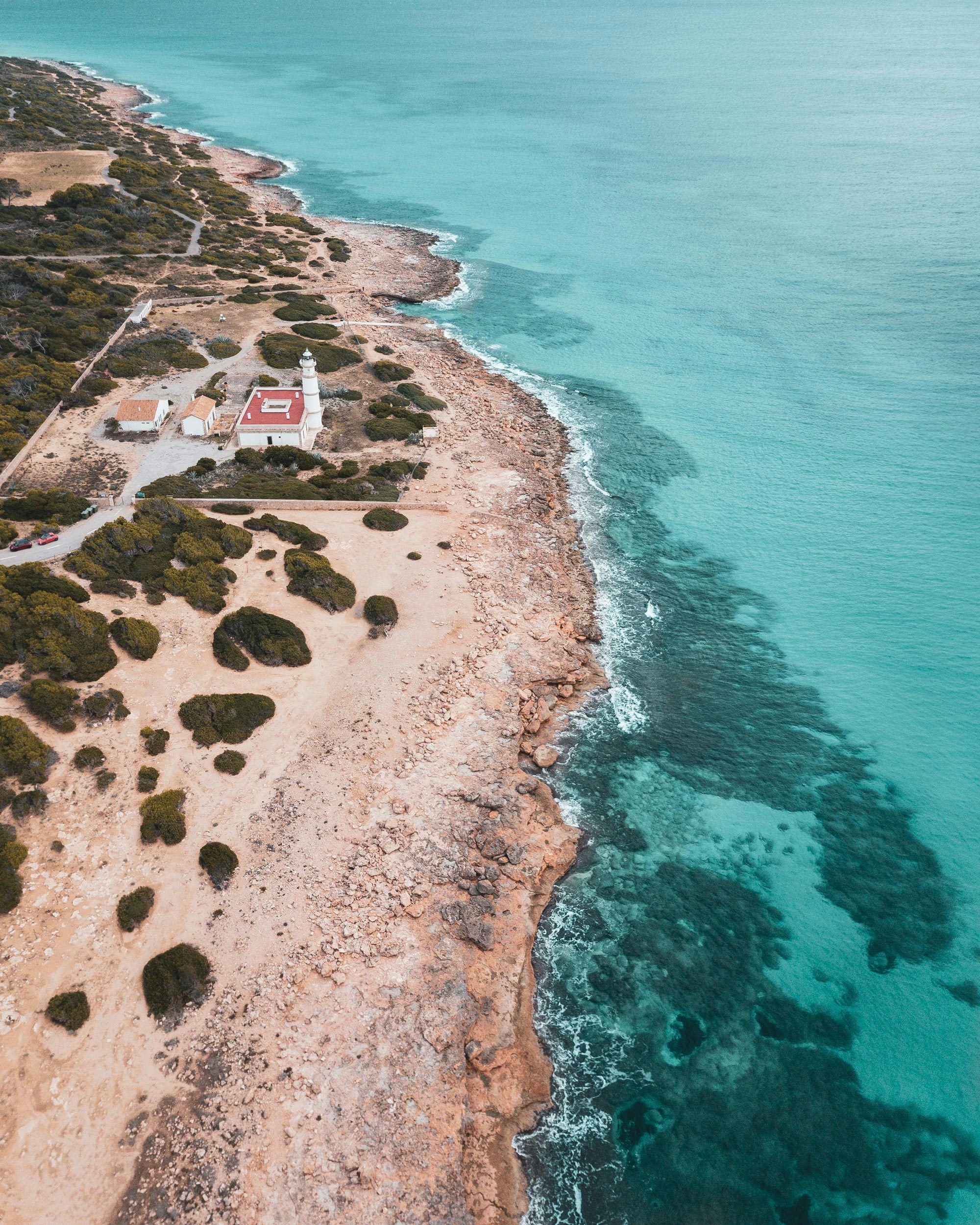 Aerial view of a lighthouse perched on Mallorca's rugged southern coastline, surrounded by turquoise waters.