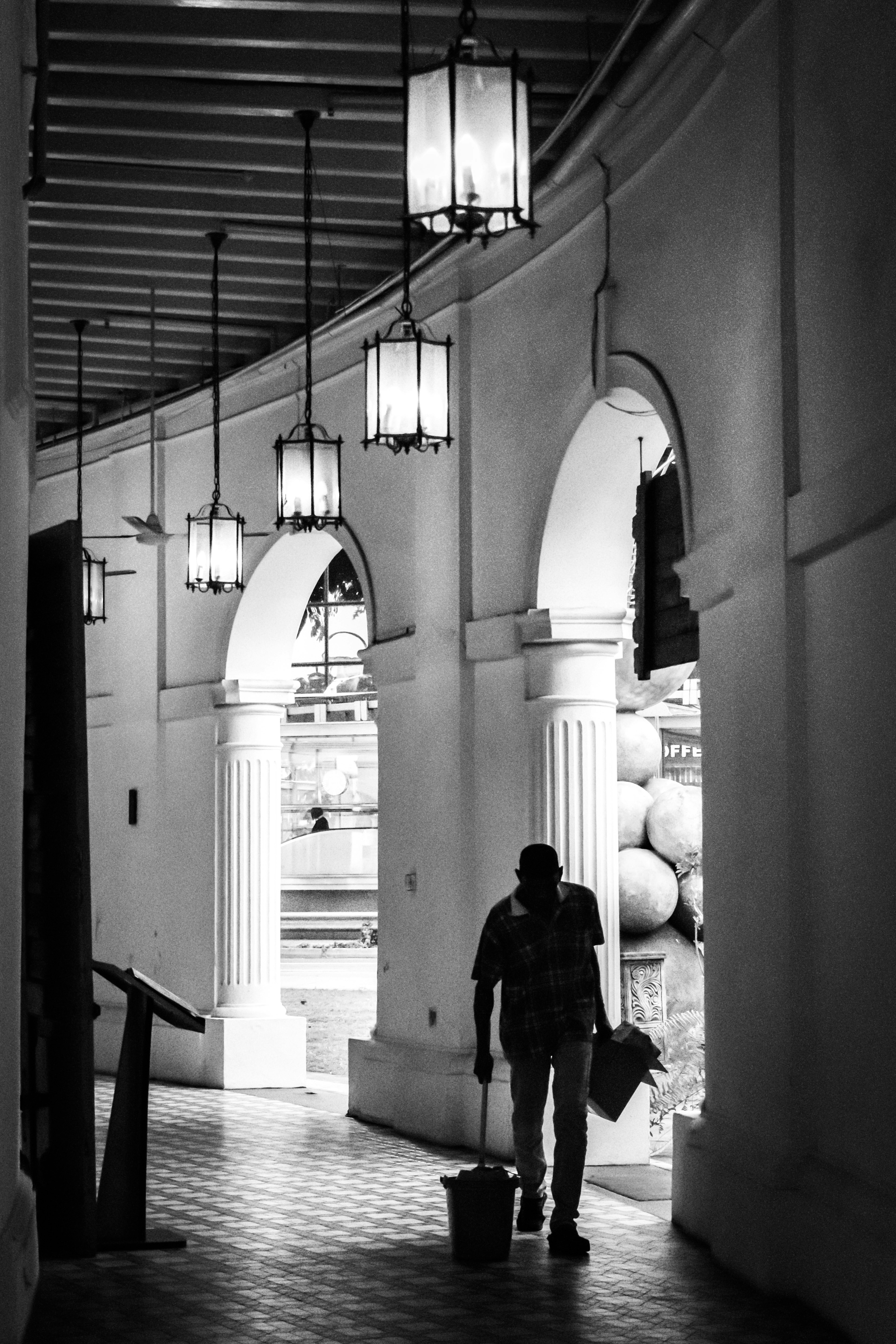 Silhouette of a man carrying a bucket in a softly lit arched corridor, framed by hanging lanterns and classical columns.