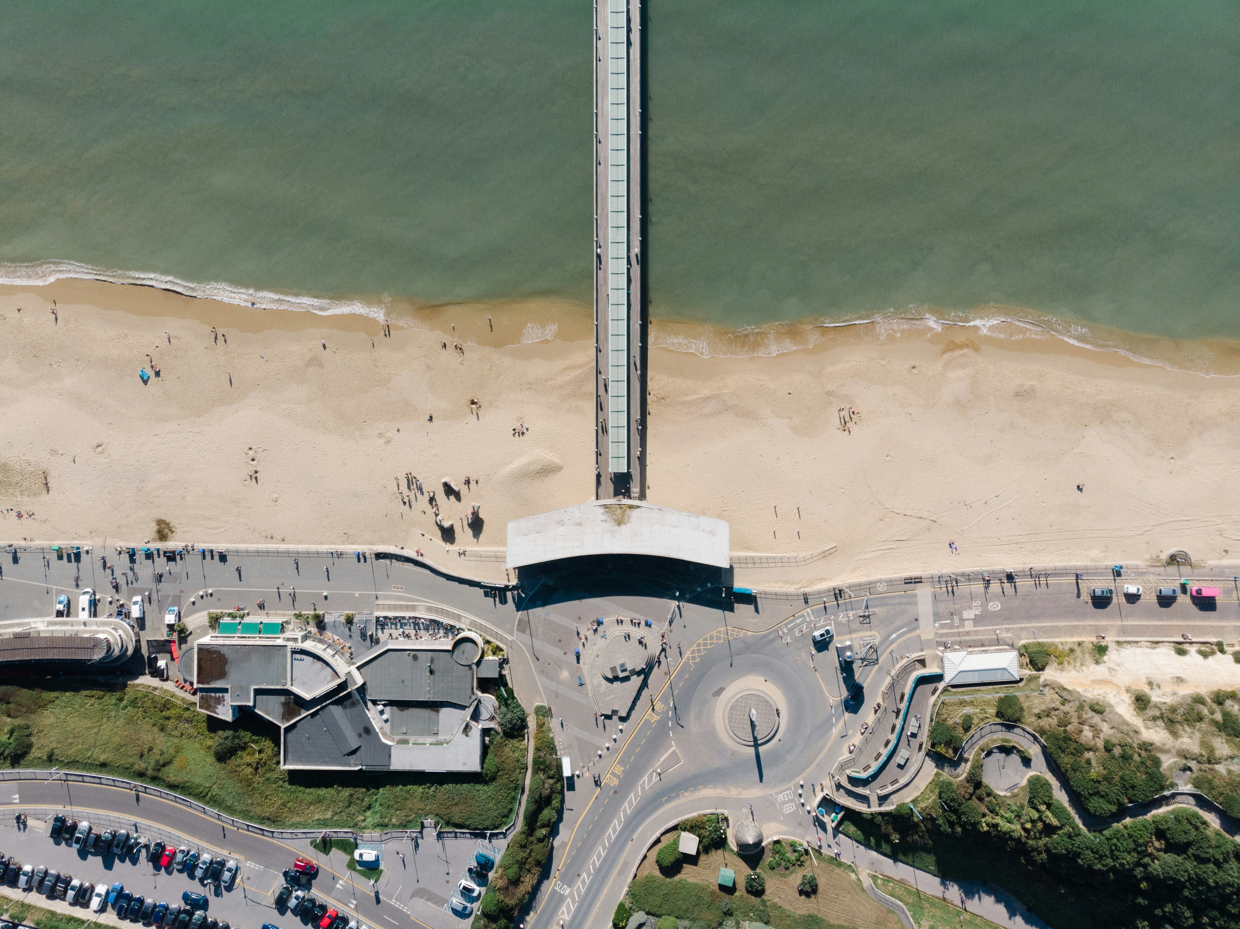 Aerial view of a pier extending into the ocean from a sandy beach, bordered by roads and buildings.