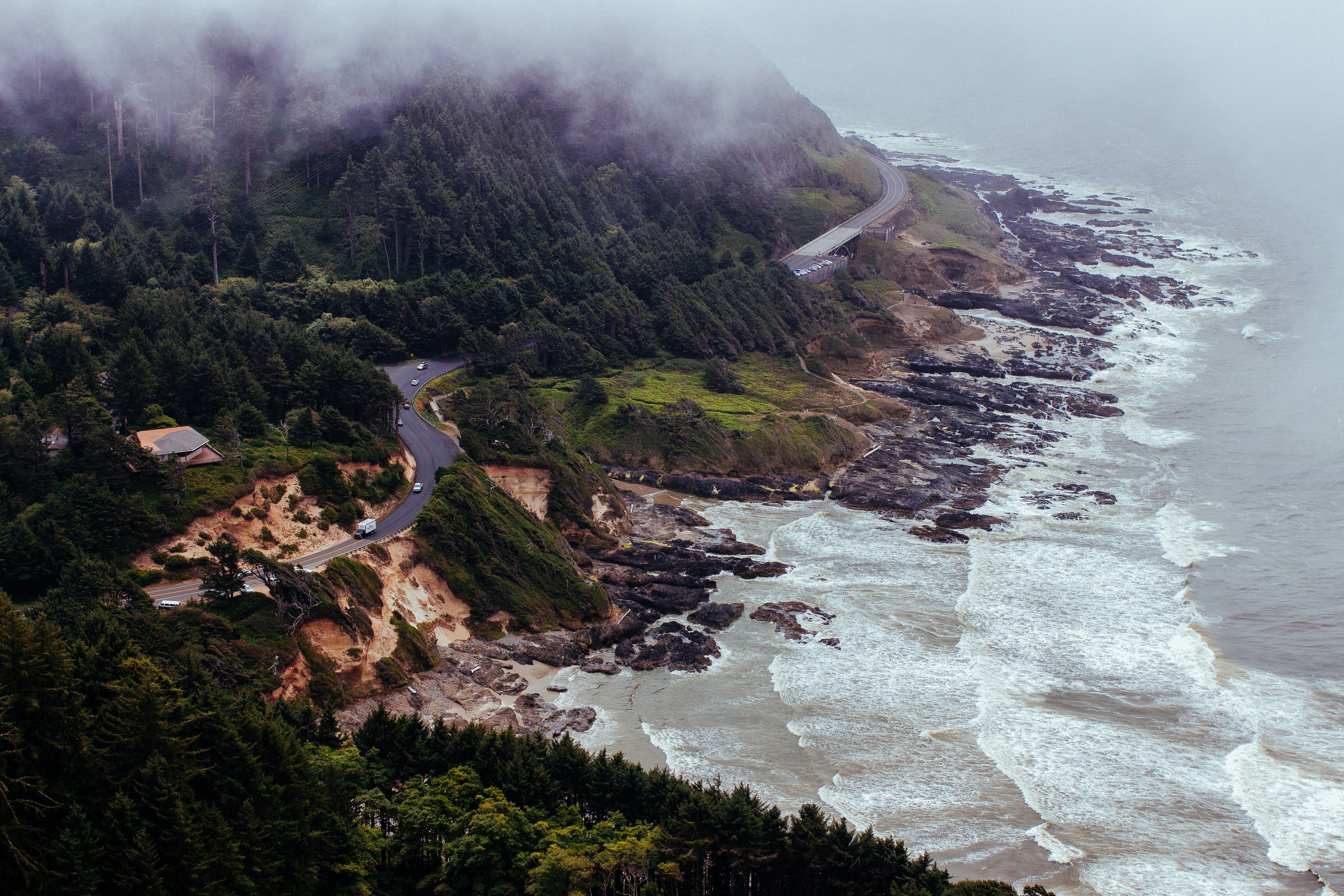 Foggy coastline with winding road and rugged cliffs meeting churning waves.