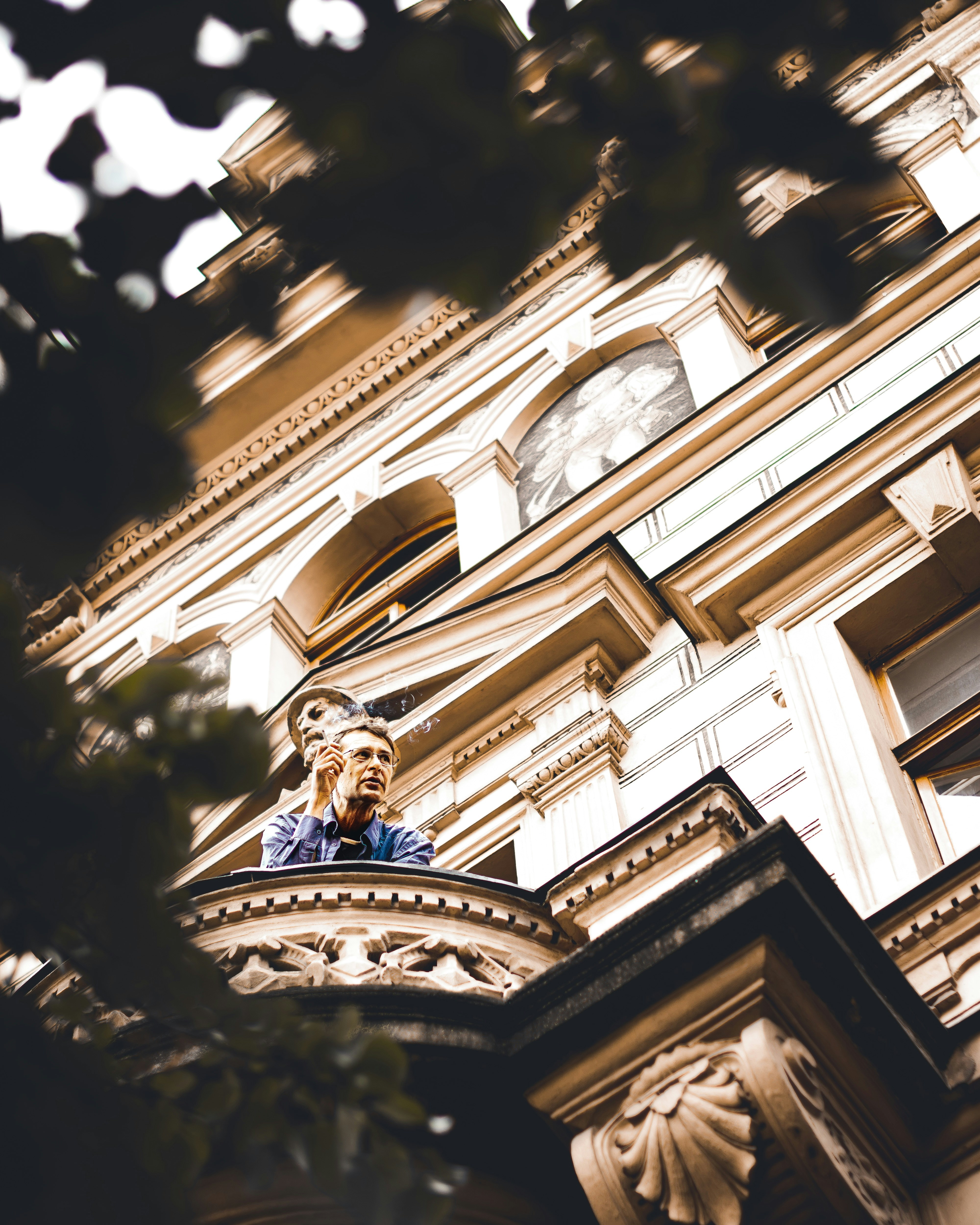 Man standing on a balcony, deep in thought, framed by lush greenery and ornate architectural details.