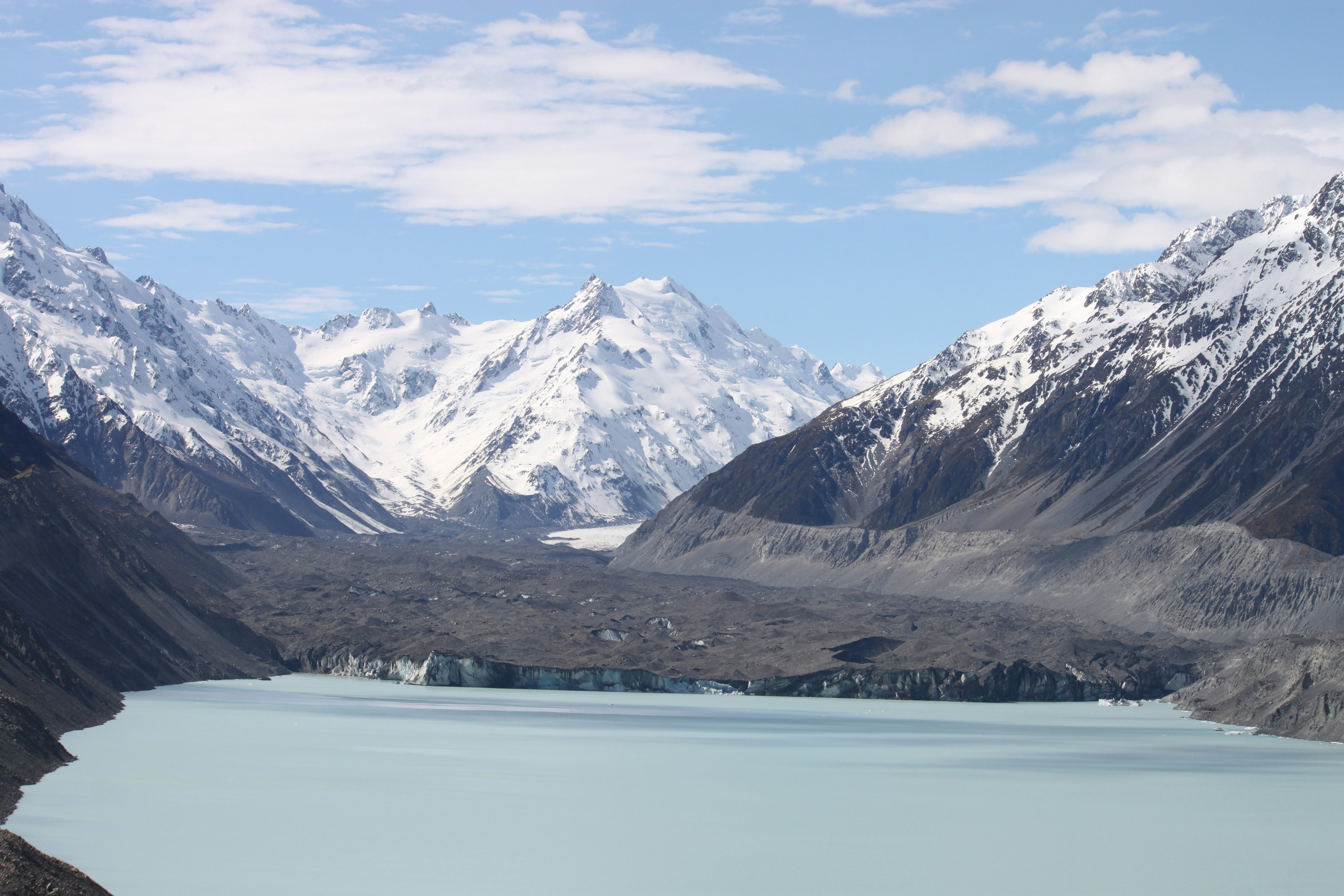 Majestic snow-capped mountains rise above a tranquil glacial lake, reflecting the clear blue sky. The scene captures the raw beauty of an untouched alpine landscape.
