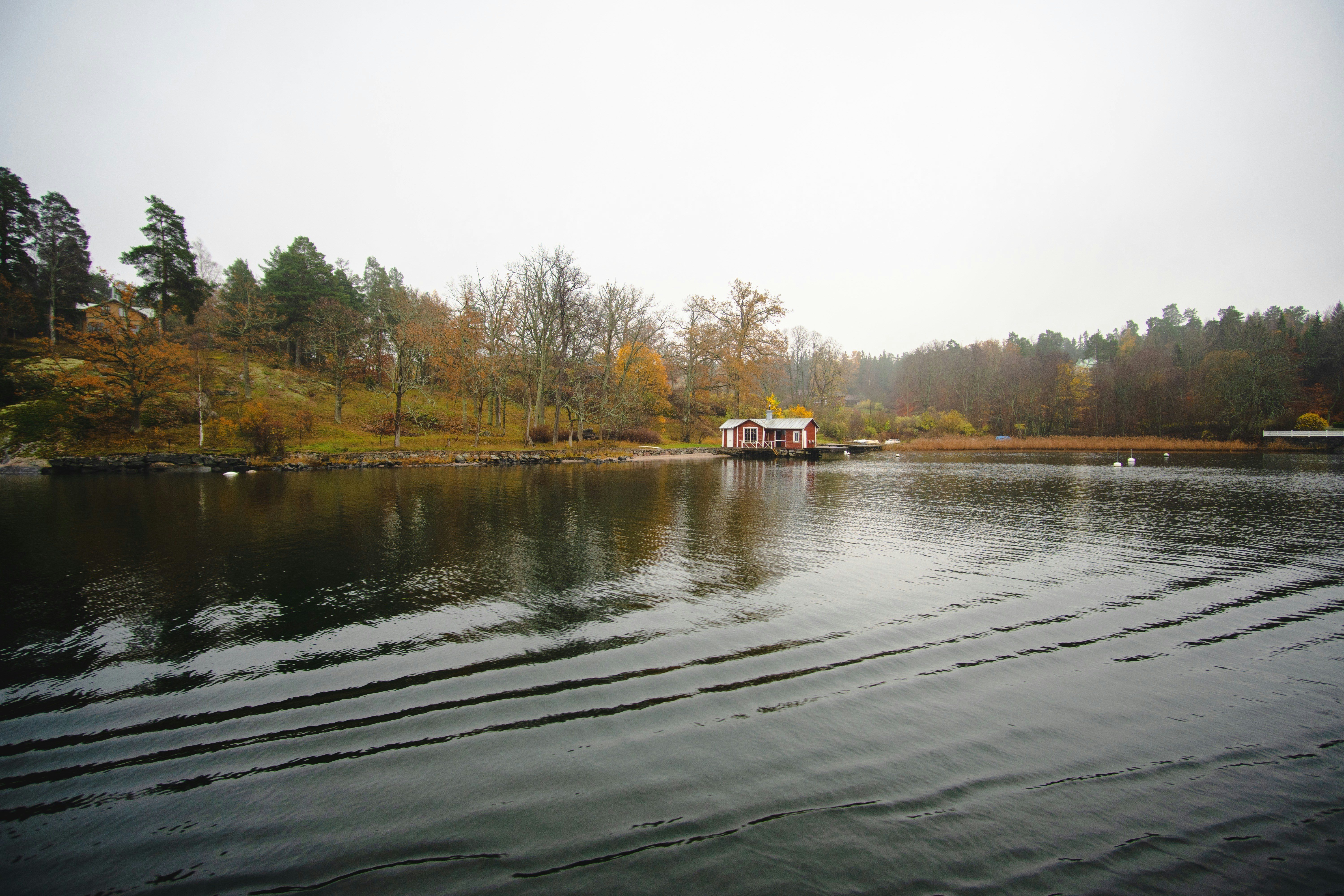 This tranquil image captures a quaint red cabin nestled on a lakeside, surrounded by autumnal trees in hues of orange, yellow, and green. The composition draws the eye across the rippling water, reflecting the overcast sky that adds a serene and peaceful atmosphere. The muted colors and soft lighting create a harmonious balance, making the scene visually striking and calming.