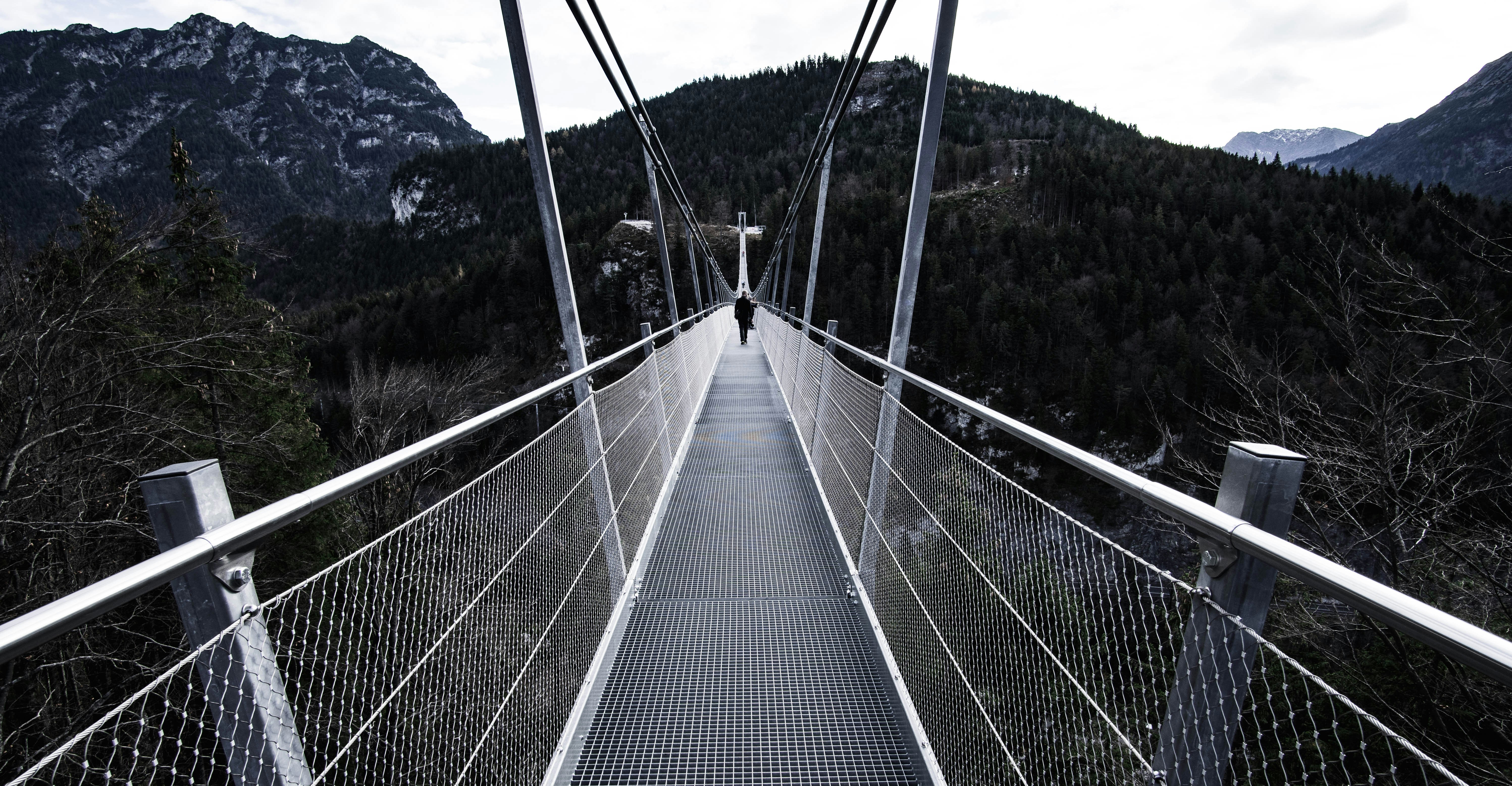 Suspension bridge stretching into a forested mountain landscape under a cloudy sky.