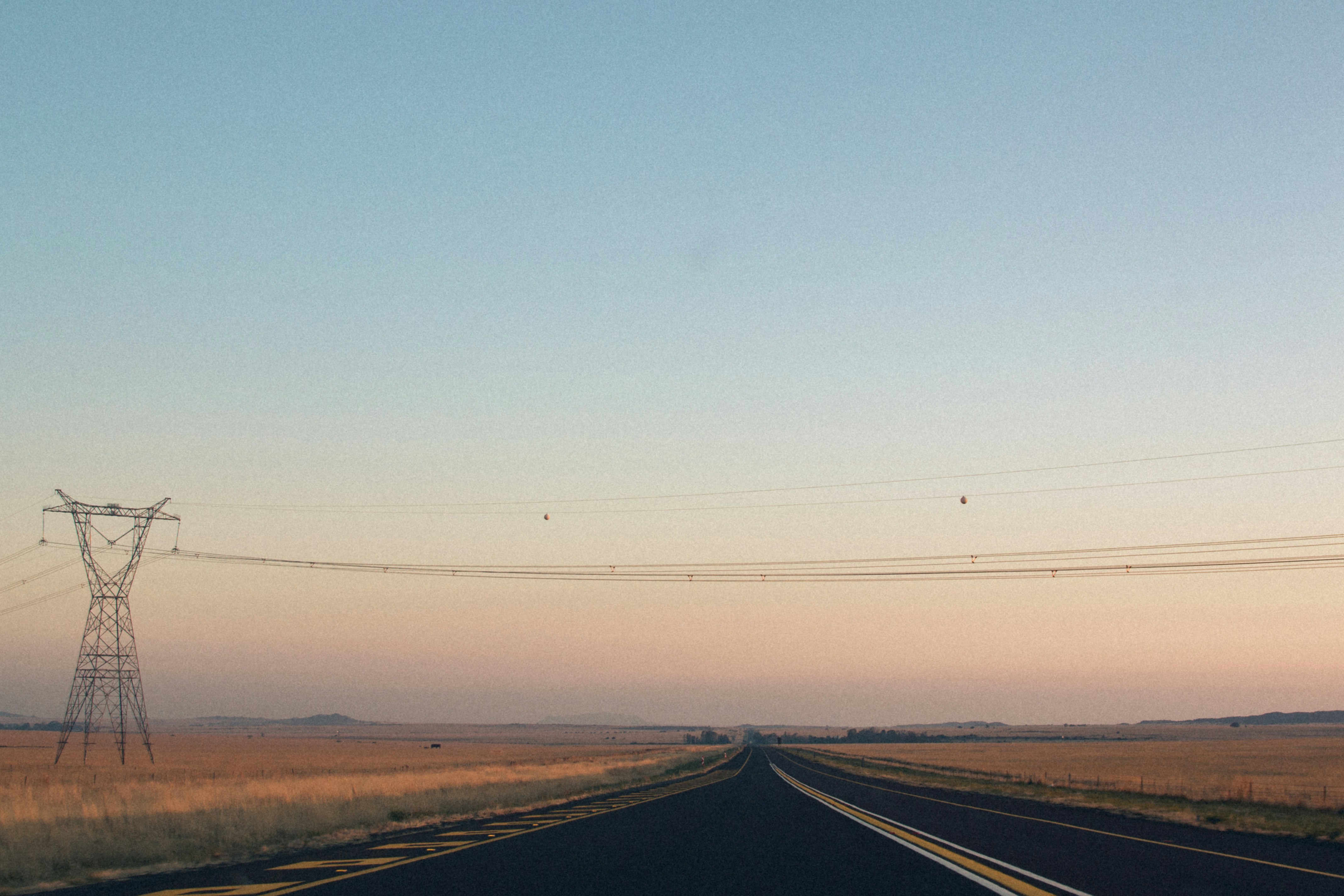 A winding road stretches into the distance, framed by golden fields and silhouetted power lines against a pastel sky.