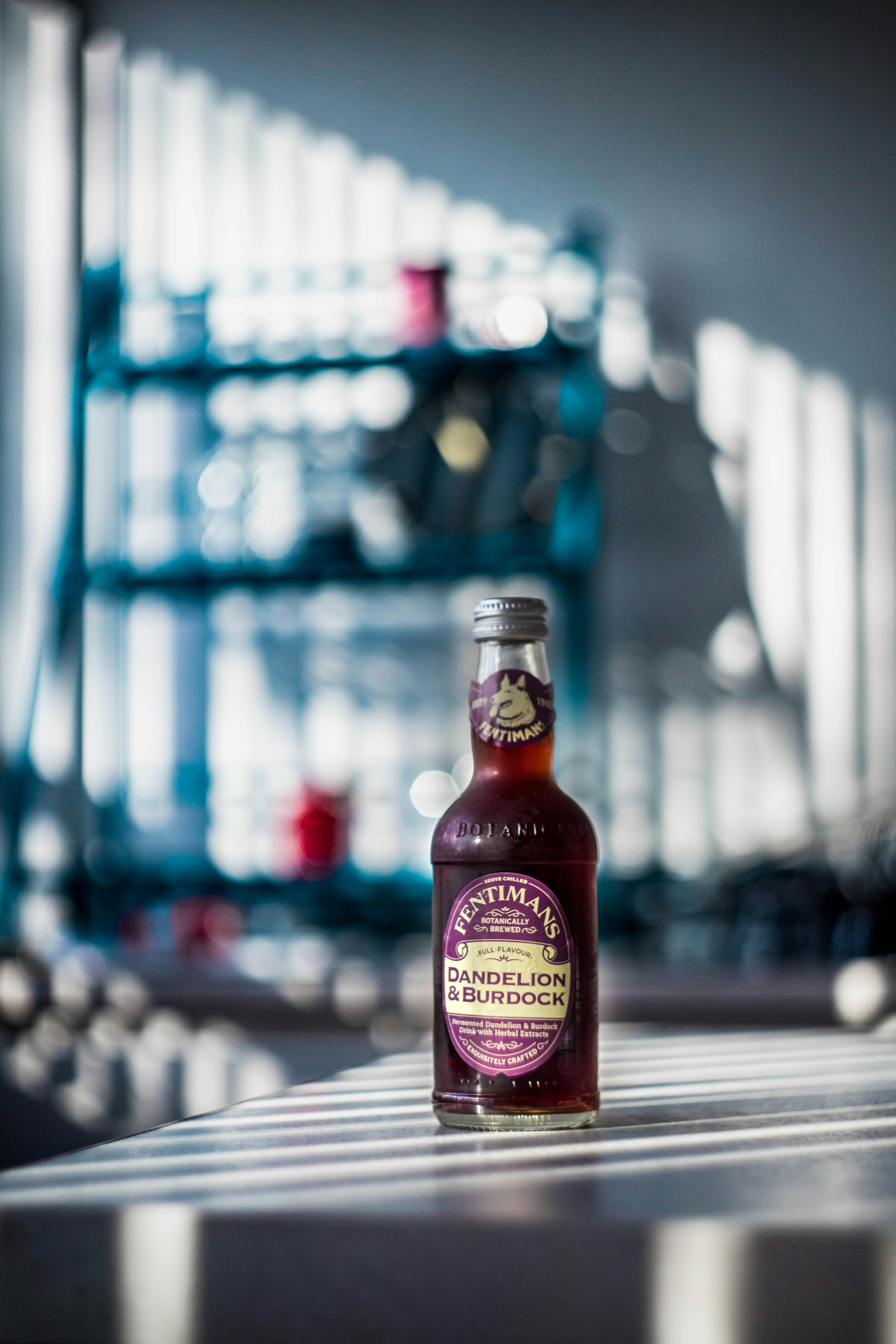 Bottle of Fentimans Dandelion & Burdock soda placed on a reflective surface, with soft shadows and blurred background elements enhancing the vintage aesthetic.