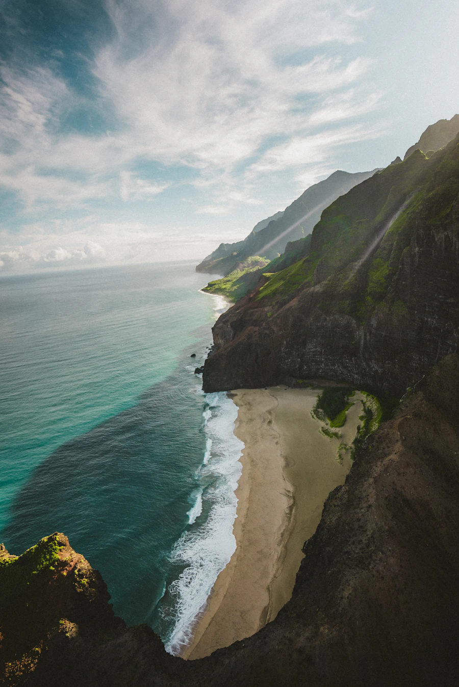 Steep green-and-black coastal cliffs overlook a narrow sandy beach and turquoise water under a bright, streaked sky.