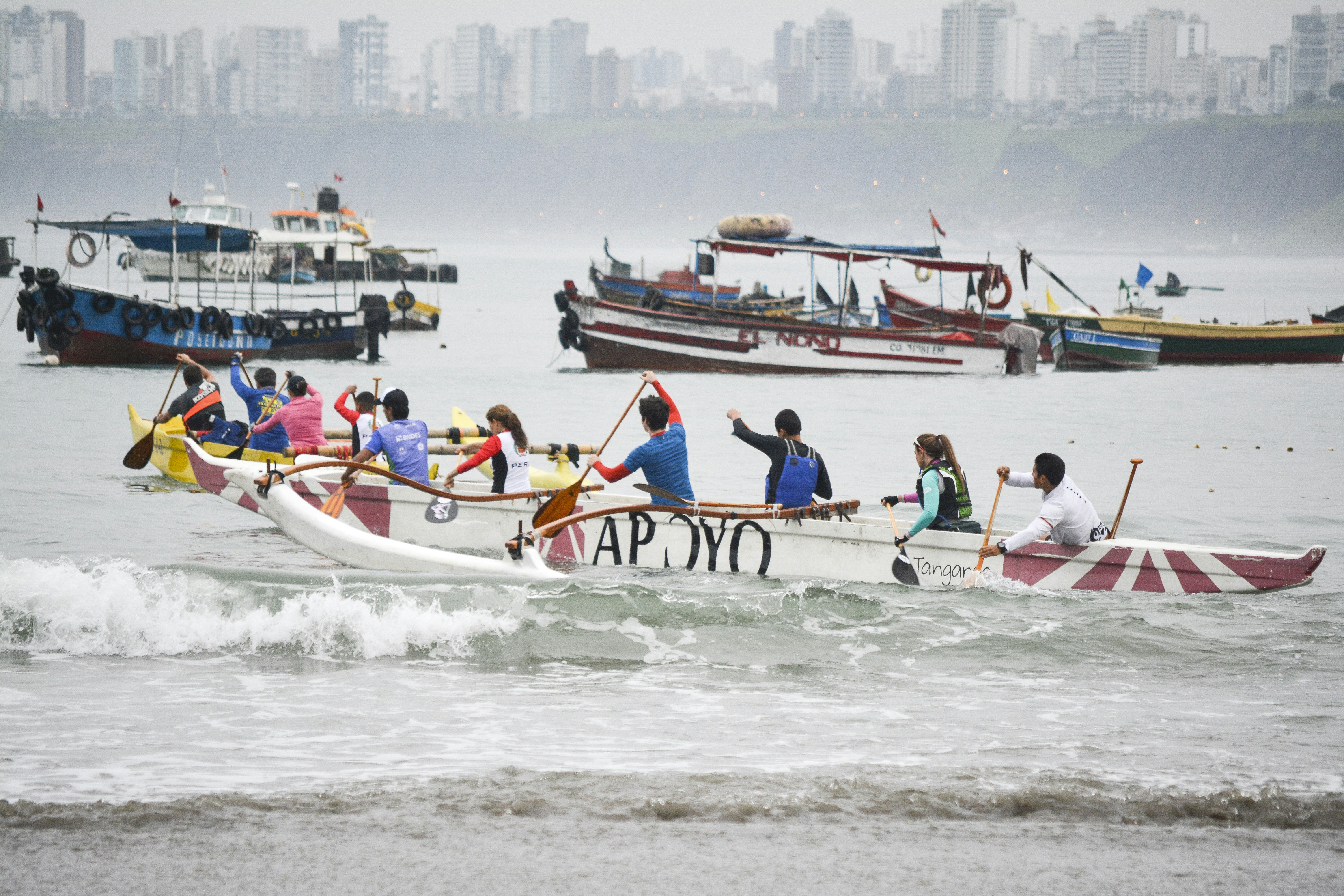 Canoe teams maneuvering through gentle waves, showcasing teamwork against a backdrop of distant city skyline and fishing boats.