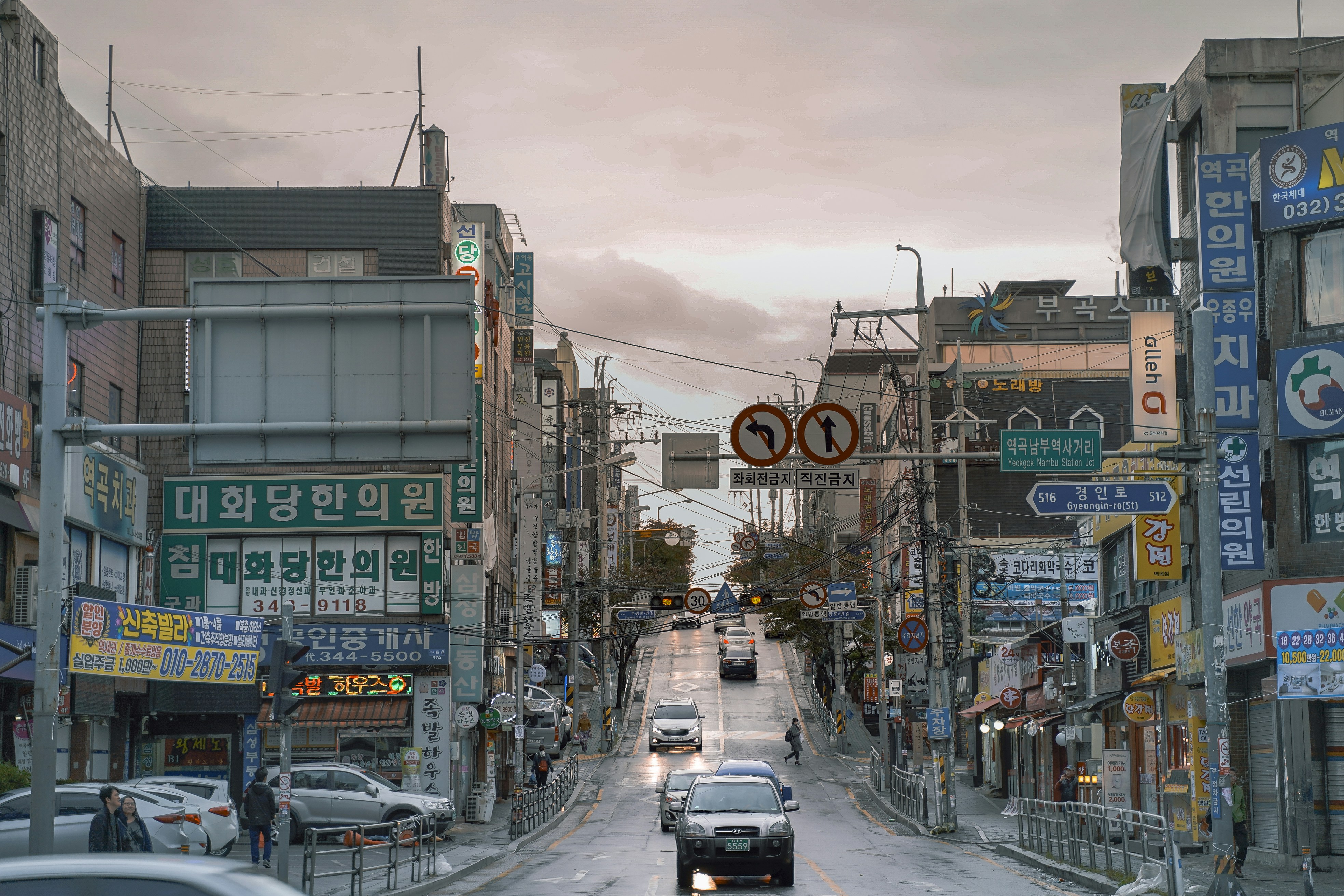 Busy street scene in a city with various storefronts and signage, capturing the everyday hustle and bustle. The road slopes gently, leading towards a series of traffic signs.