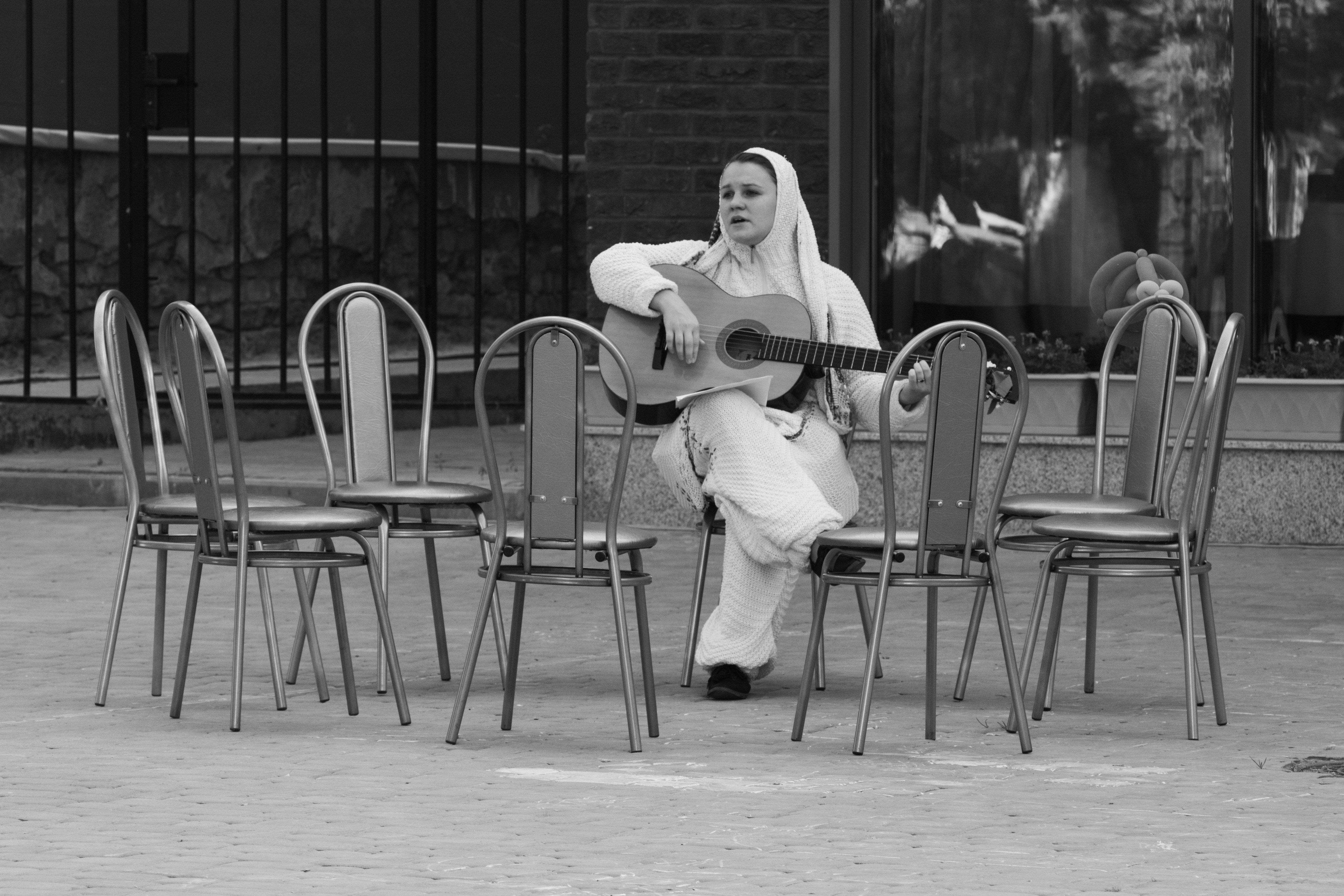 Black and white scene of a woman playing guitar while seated among empty chairs in an outdoor setting.