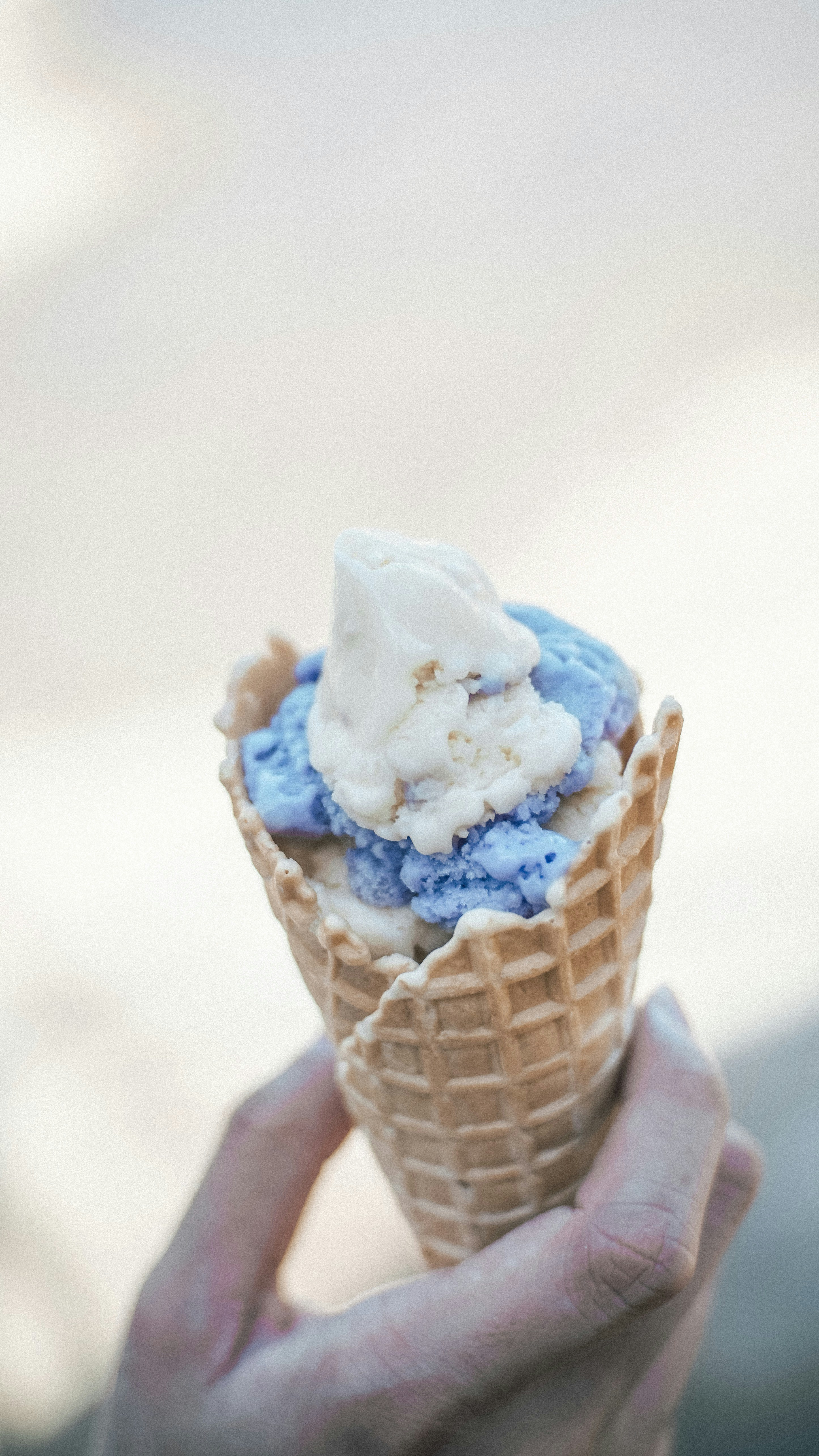An ice cream cone featuring a blend of blue and white flavors held by a hand. The soft focus background enhances the treat's vibrant colors.
