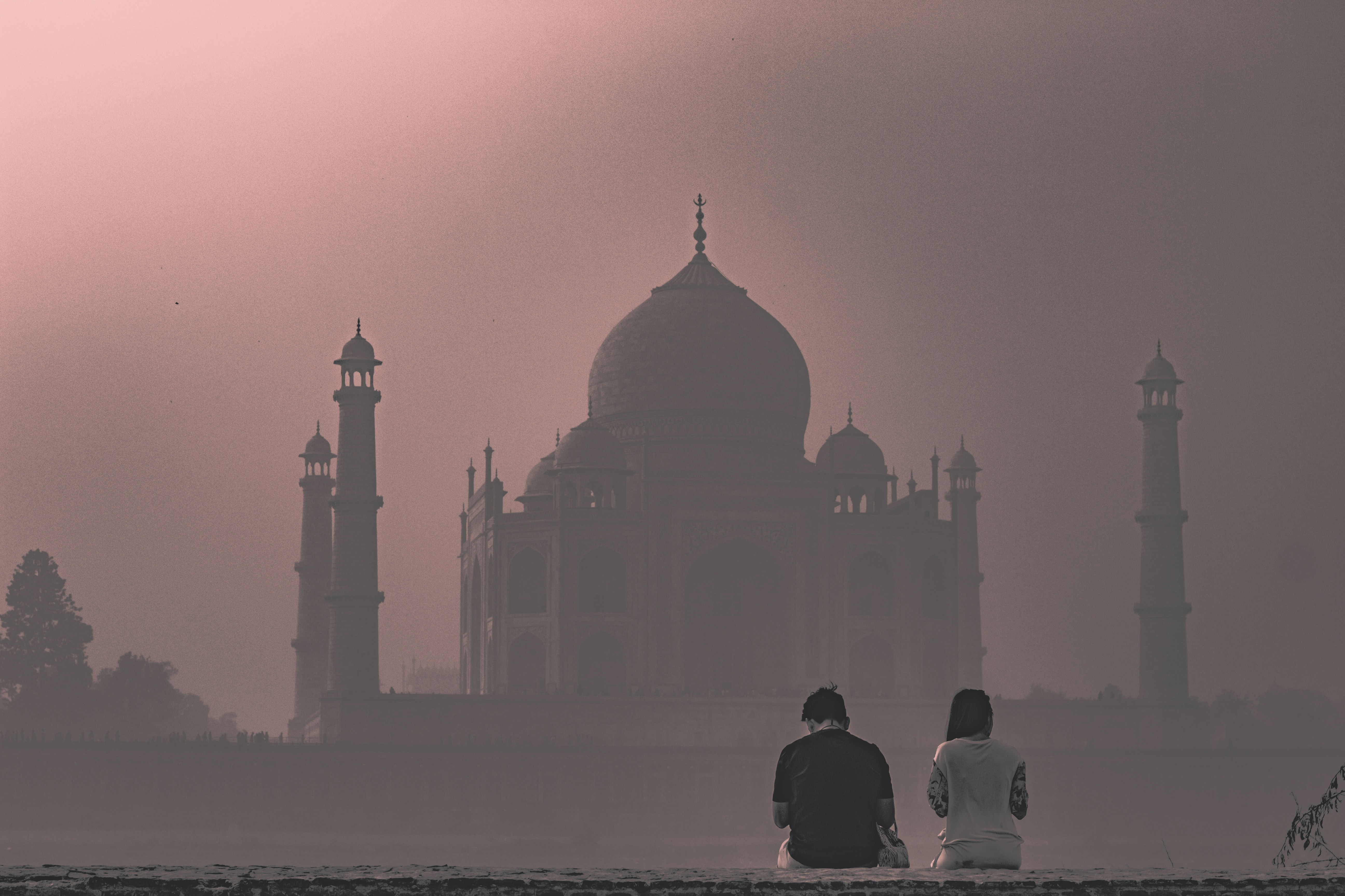 Silhouetted figures seated in front of the Taj Mahal at sunrise, shrouded in a soft pink mist.