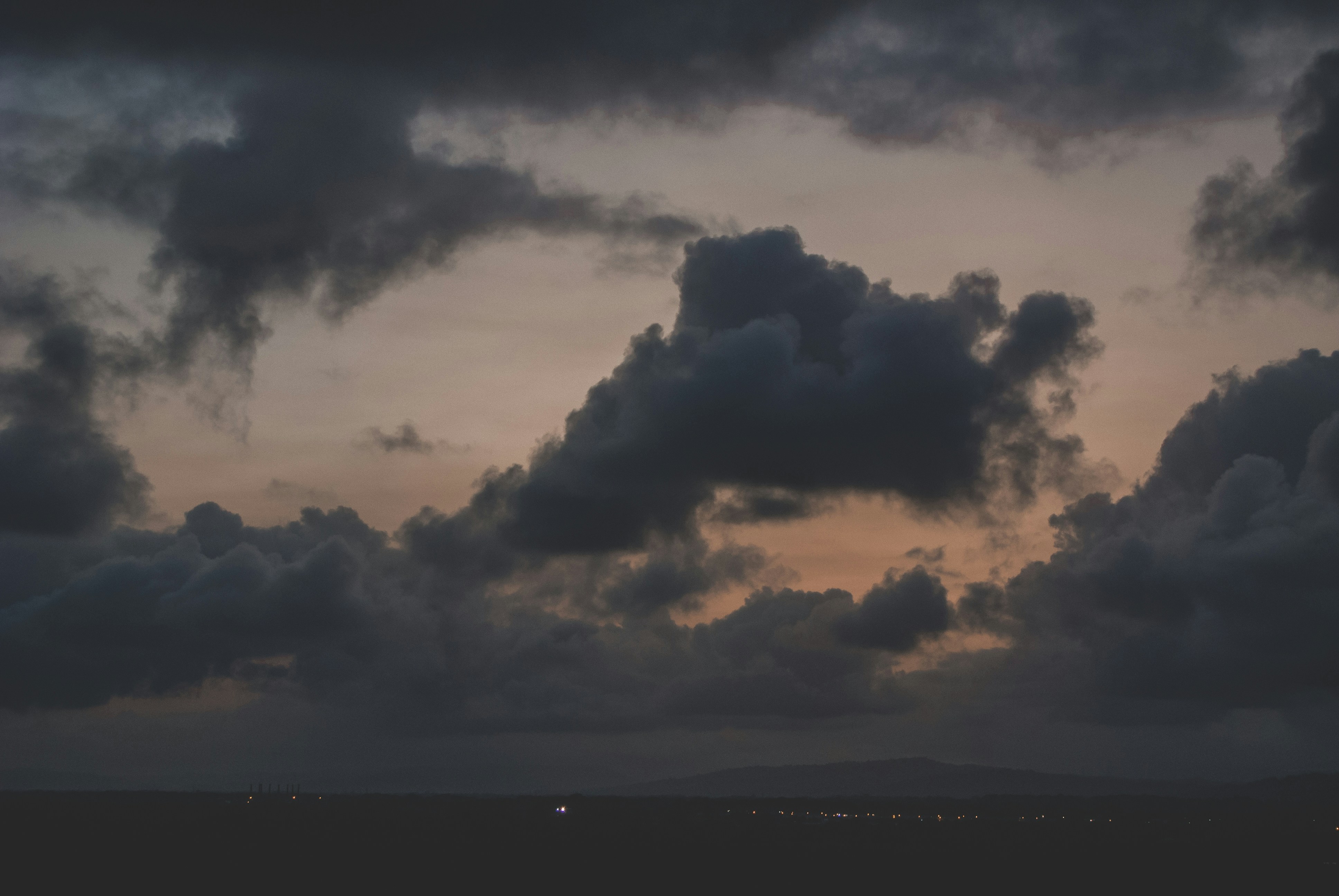 Cloud-laden dusk sky with a warm orange glow along the horizon over a dark, calm sea. Distant coastline lights punctuate the silhouette.