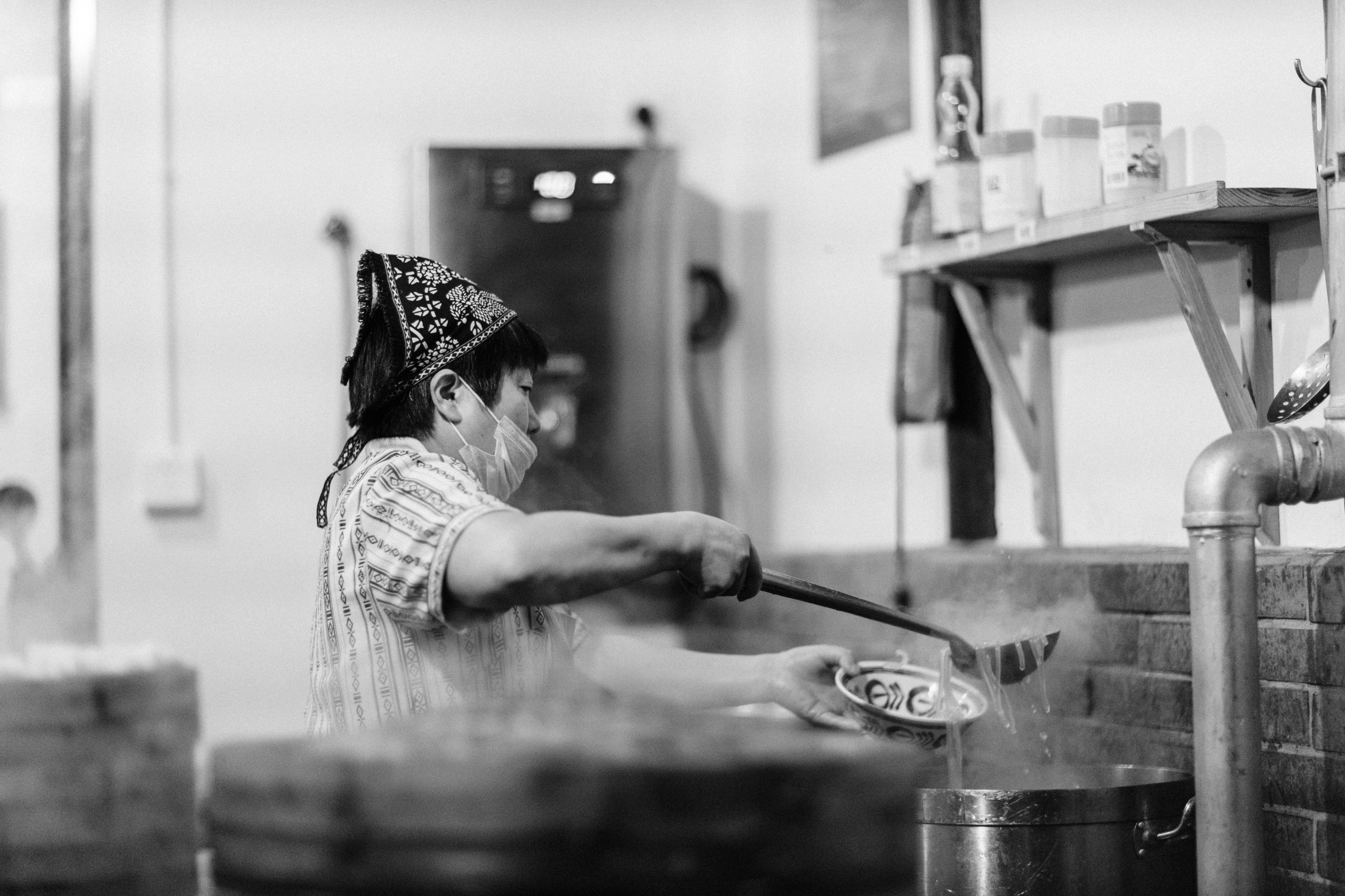 Woman wearing a patterned headscarf and mask serves steaming noodles from a pot in a rustic kitchen.