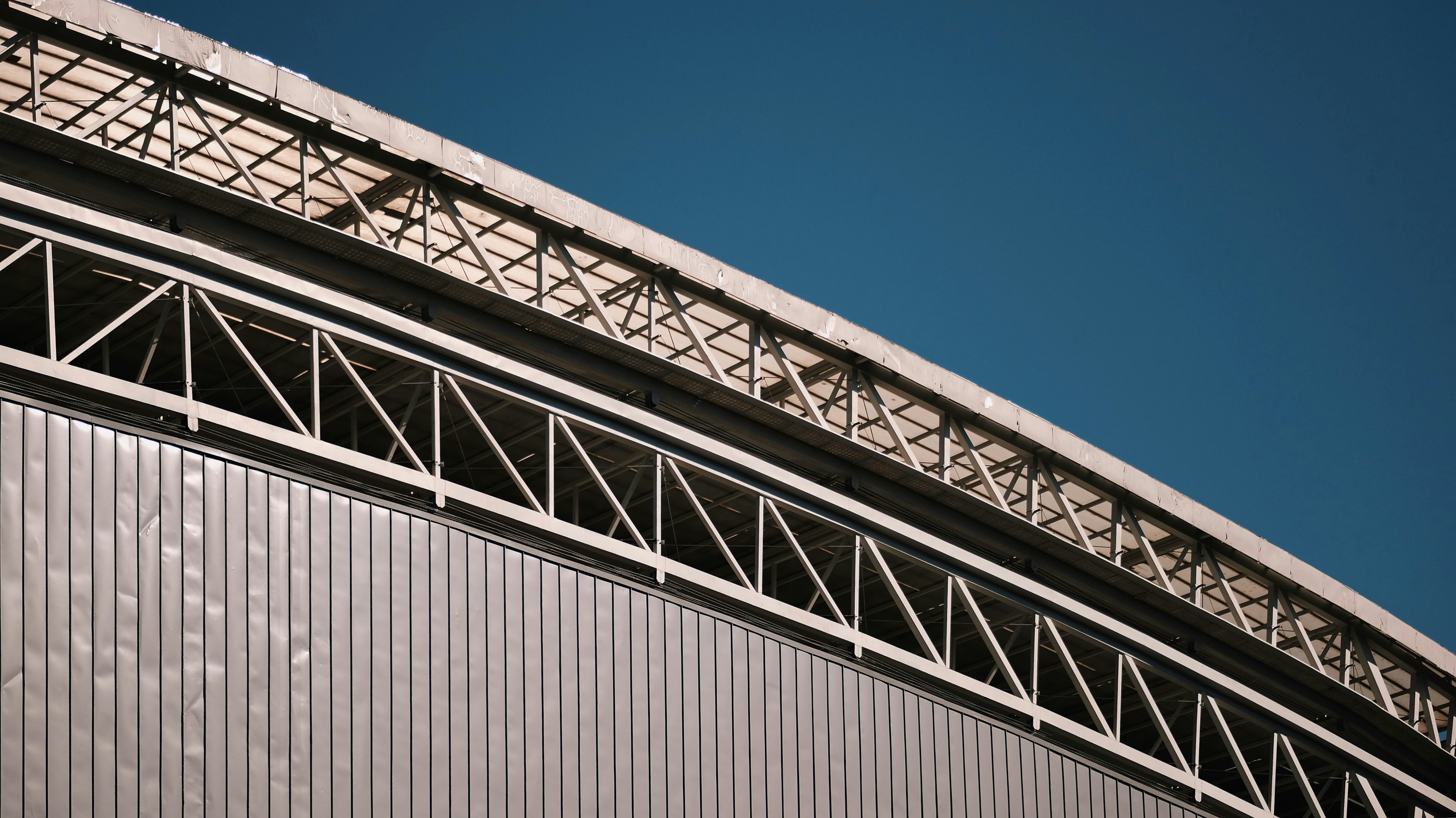 This striking image captures the sweeping curve of a modern architectural structure against a deep blue sky. The composition highlights the intricate lattice of metal beams, creating a dynamic interplay of lines and shadows. The warm tones of the building contrast beautifully with the crisp, clear sky, emphasizing the structure's geometric elegance and the serene atmosphere of the scene.